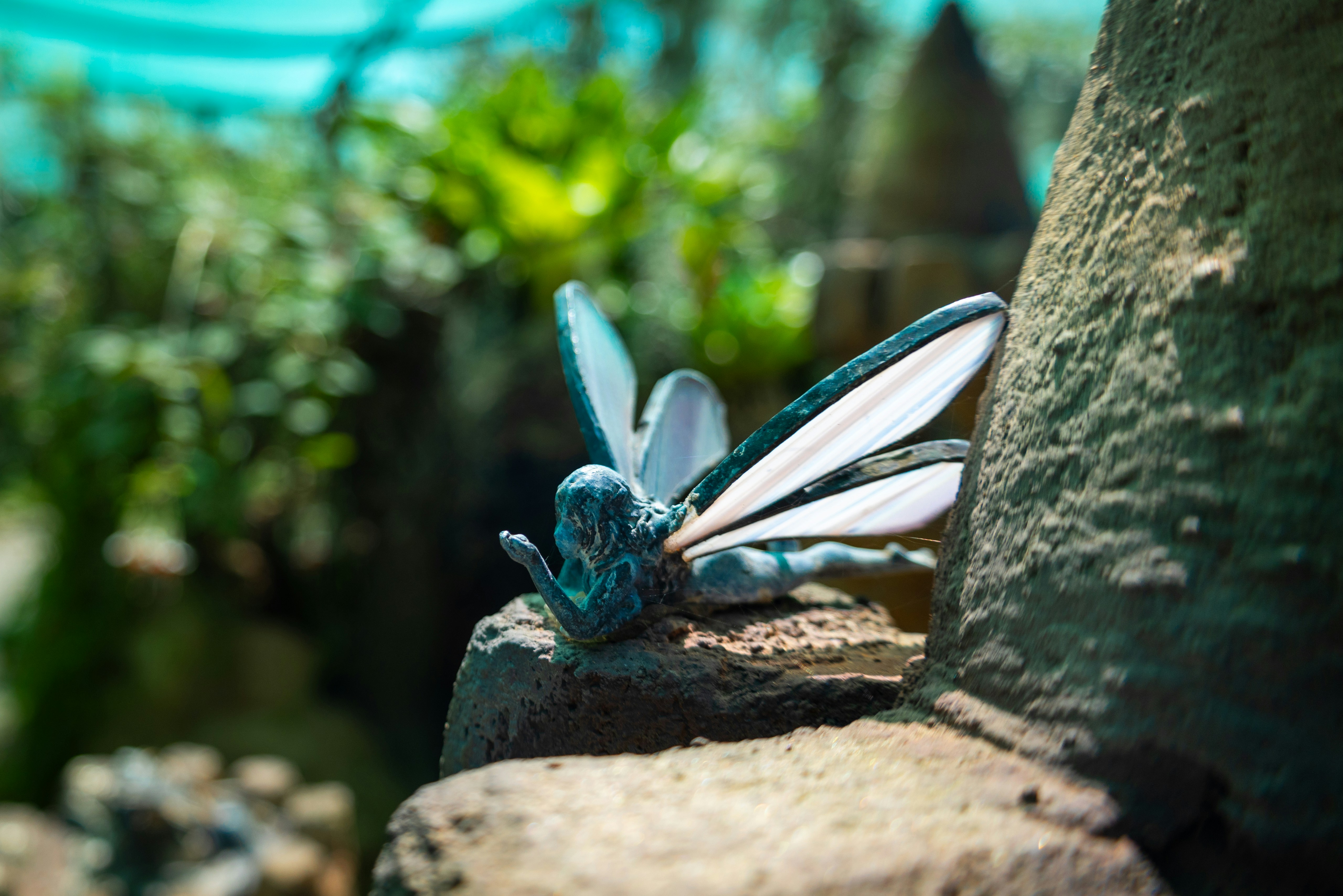 A whimsical fairy figurine perched on a stone, surrounded by lush greenery and soft light filtering through foliage.