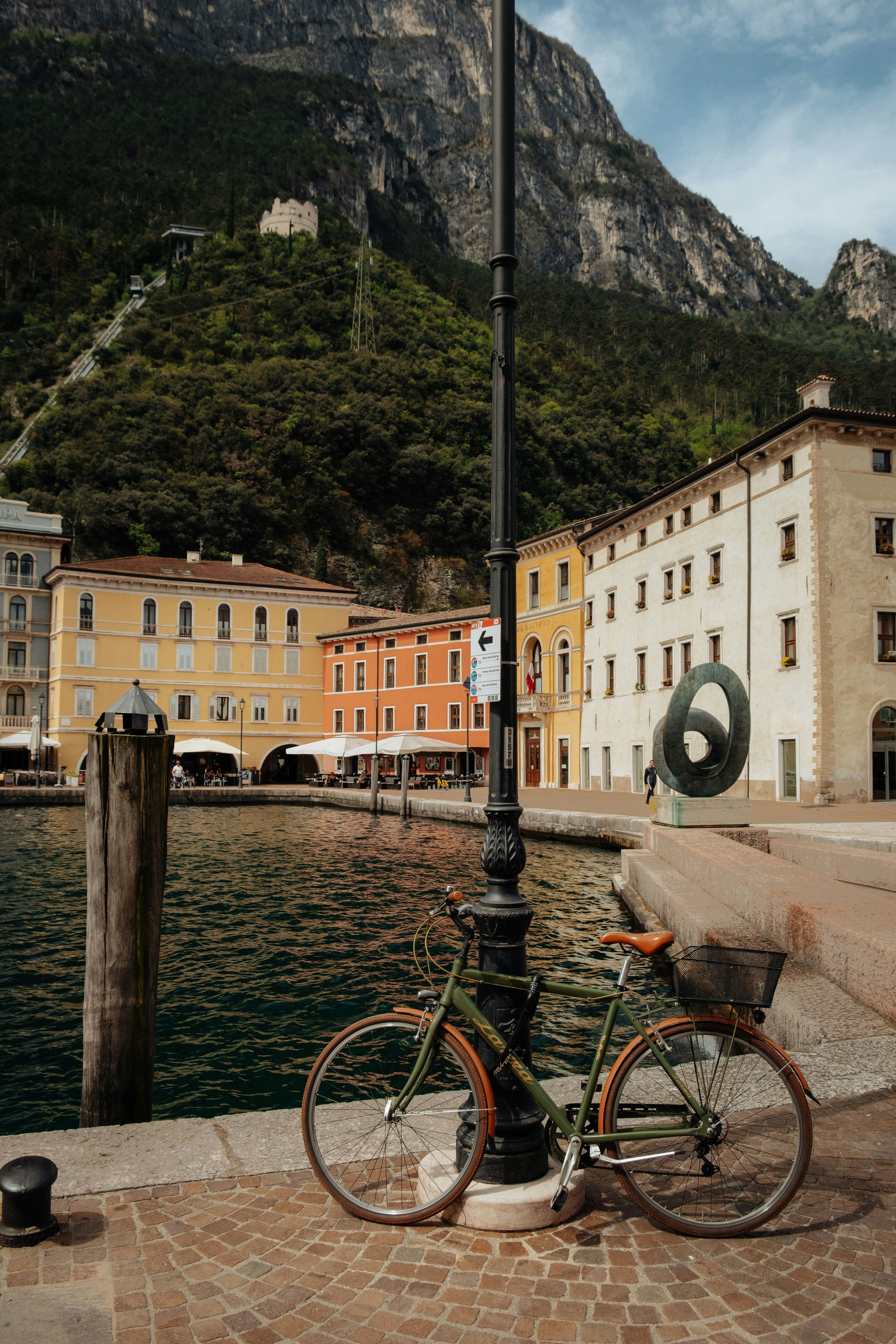 A vintage bicycle rests against a lamppost by the tranquil waters of Lake Garda, framed by colorful buildings and lush mountains in the background.