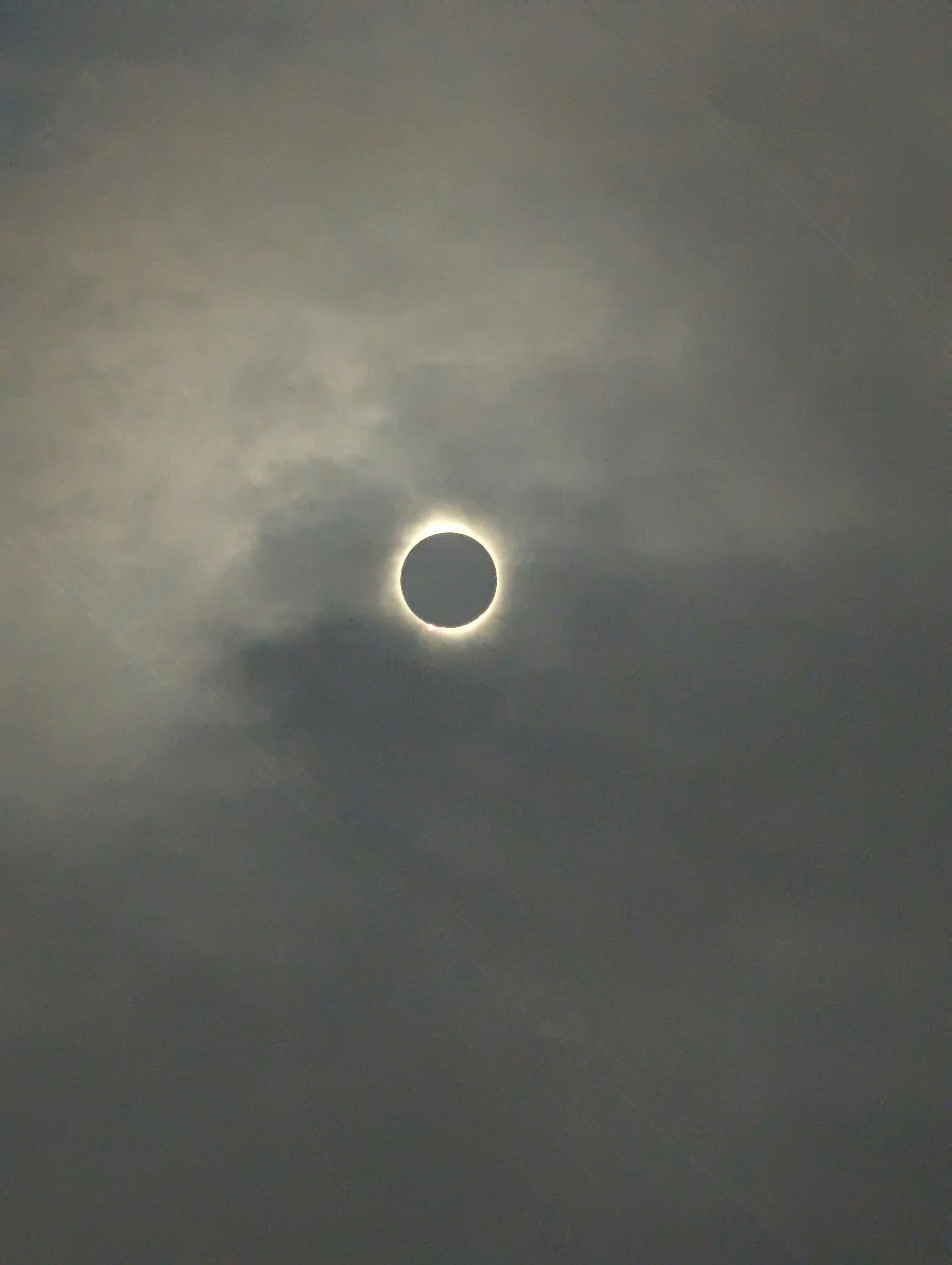 Photograph capturing a solar eclipse where the Moon obscures the Sun, revealing a delicate corona. Clouds drift across a dim sky, emphasizing the circular halo around the solar disk.