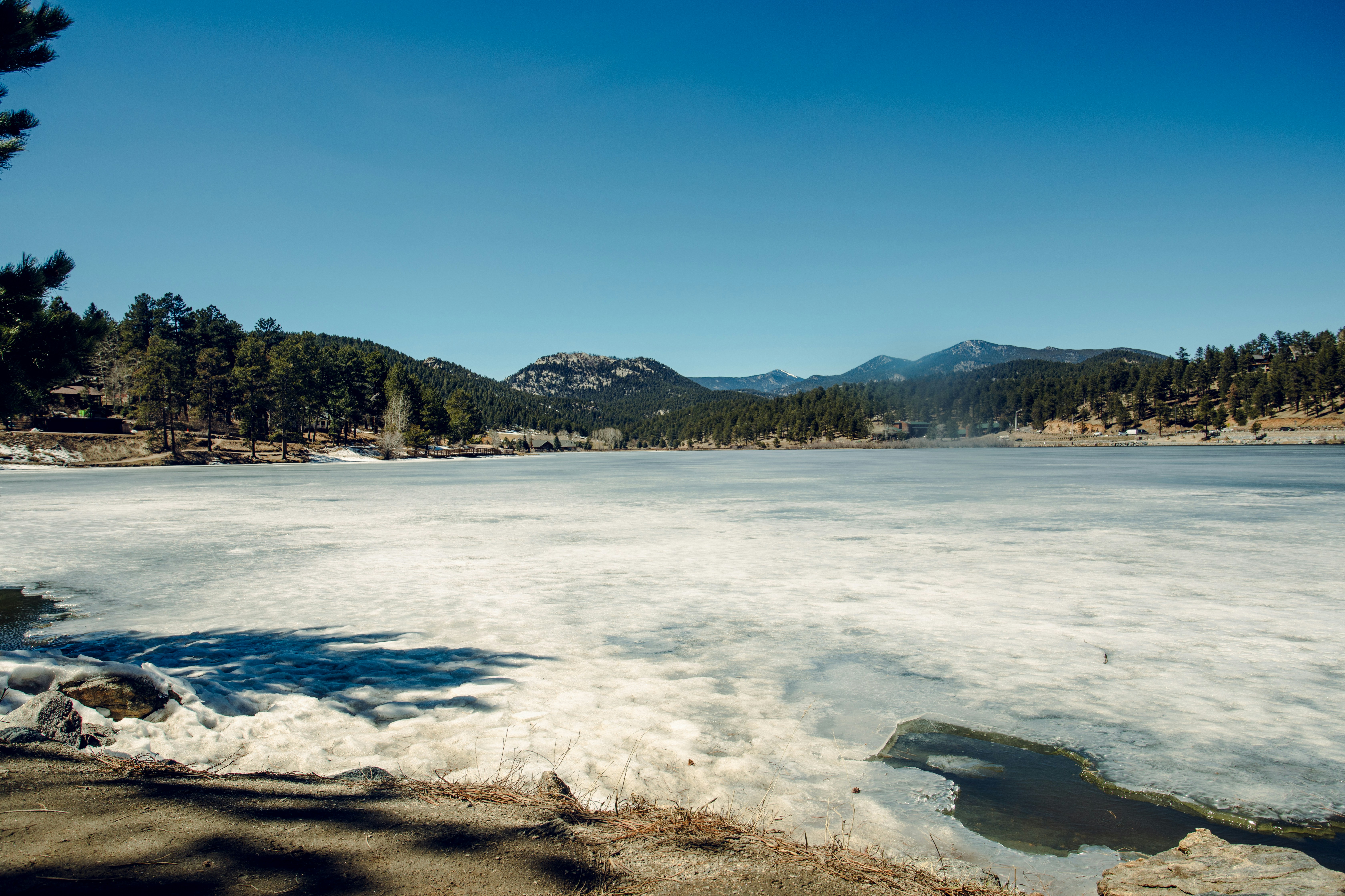 A serene frozen lake surrounded by evergreen trees and distant mountains under a clear blue sky.