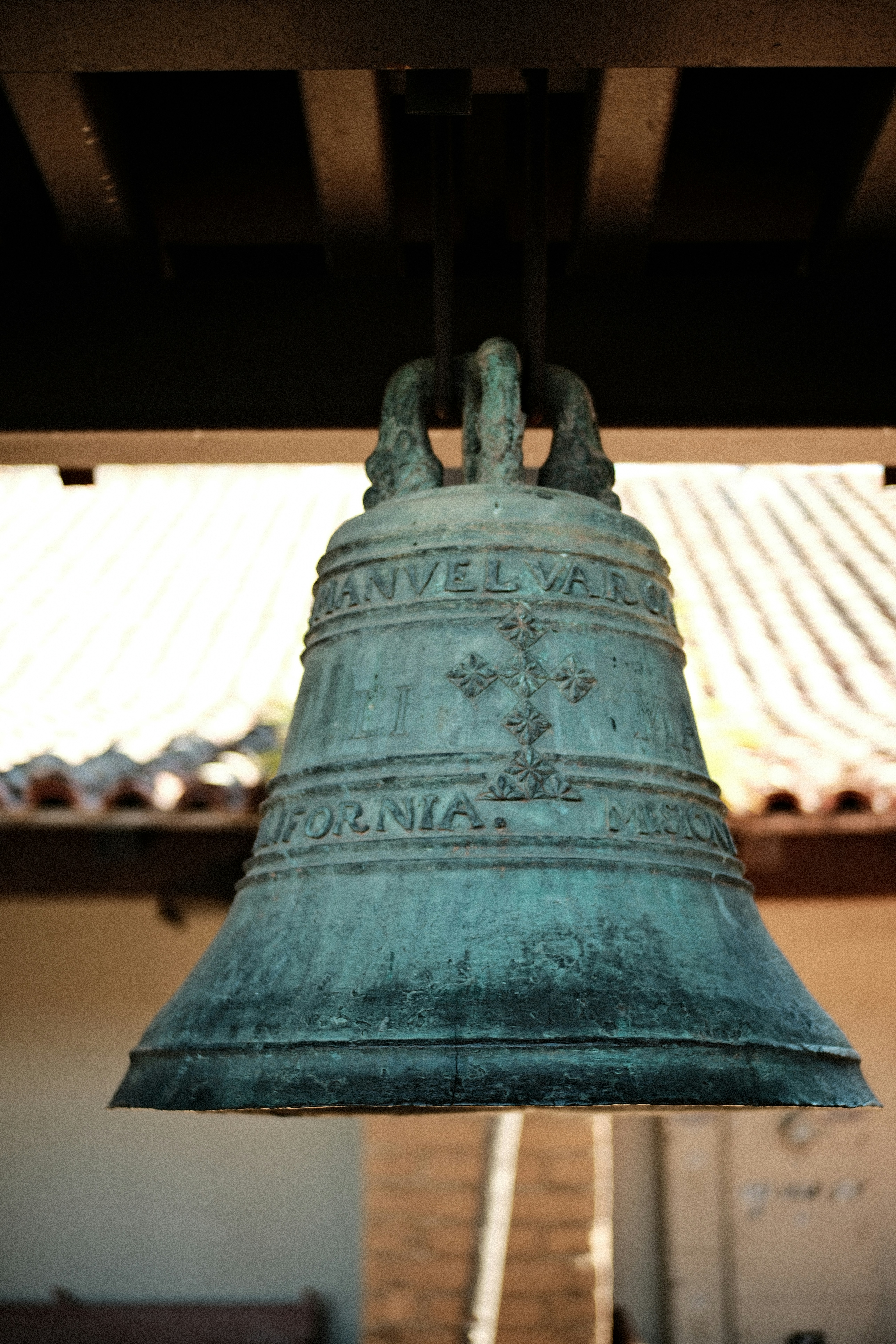 Patinated Bronze Bell Under Wooden Beams in a Sunlit Courtyard