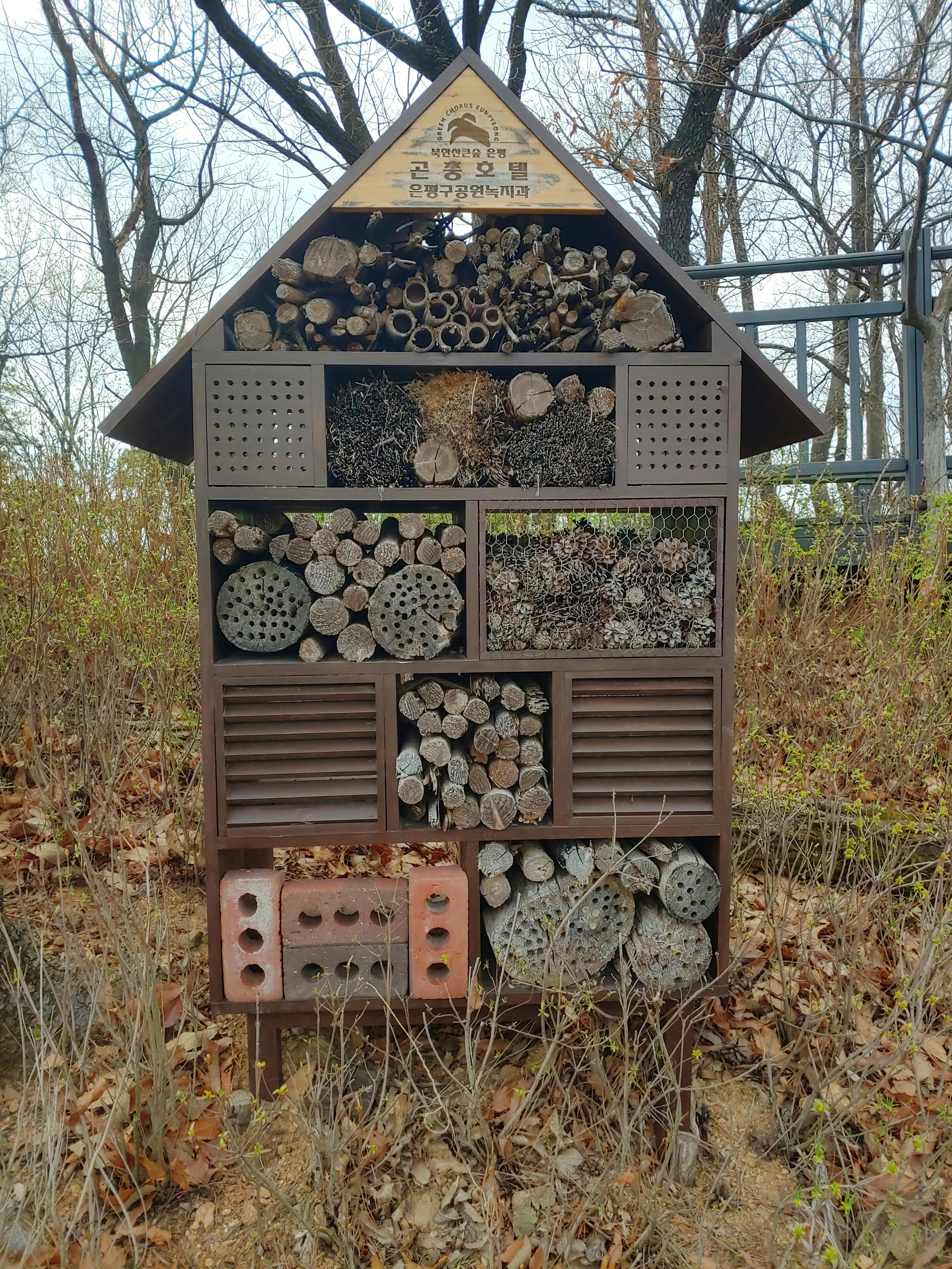 A multi-compartment wooden insect hotel filled with hollow logs and drilled tubes sits in a leaf-strewn clearing. The structure serves as a habitat for pollinators and beneficial insects.