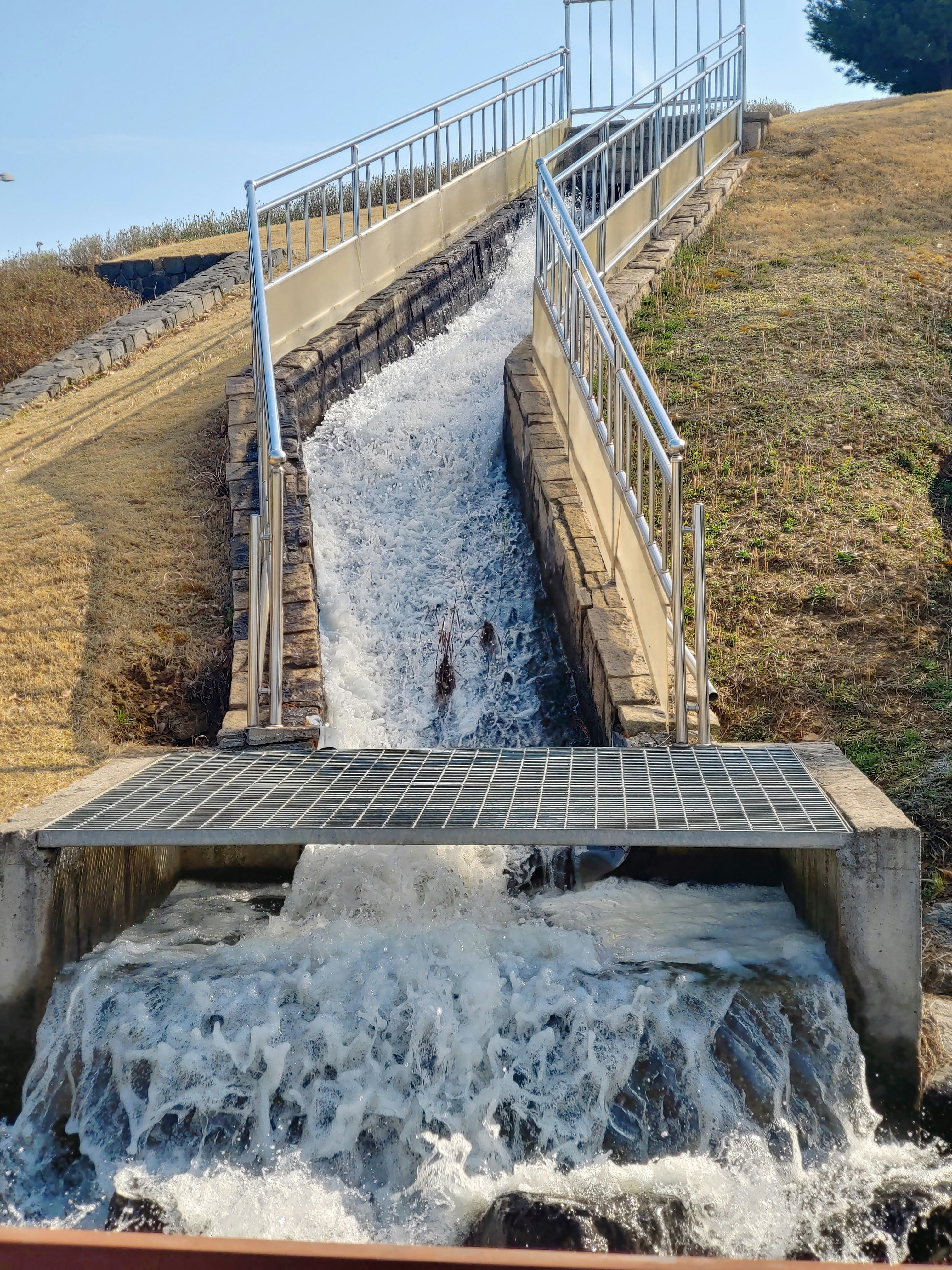 Concrete spillway channels a torrent down a stepped bed, framed by metal railings and grassy slopes under bright daylight.