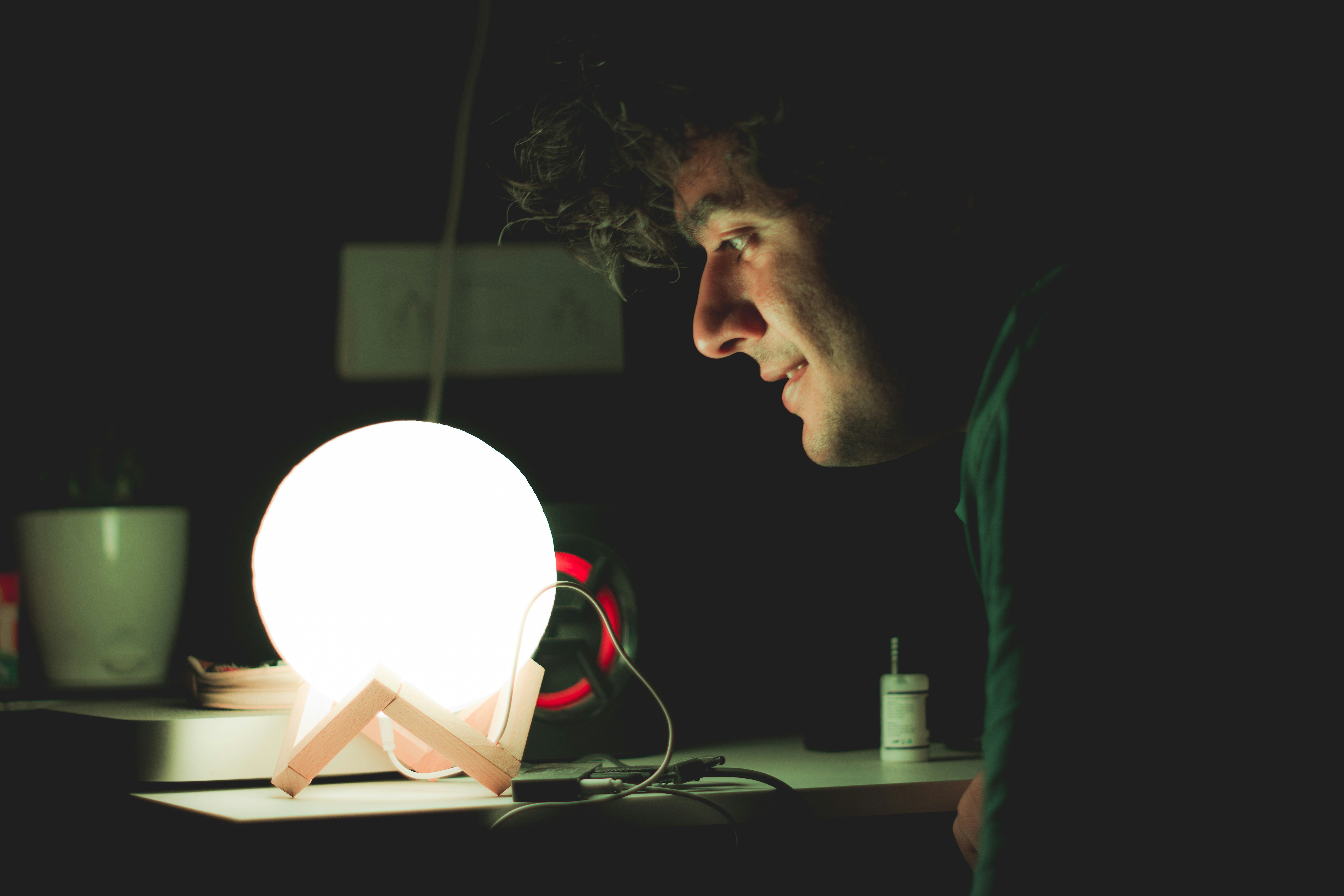 A man gazes intently at a glowing spherical lamp on a dimly lit table, highlighting the interplay of light and shadow.