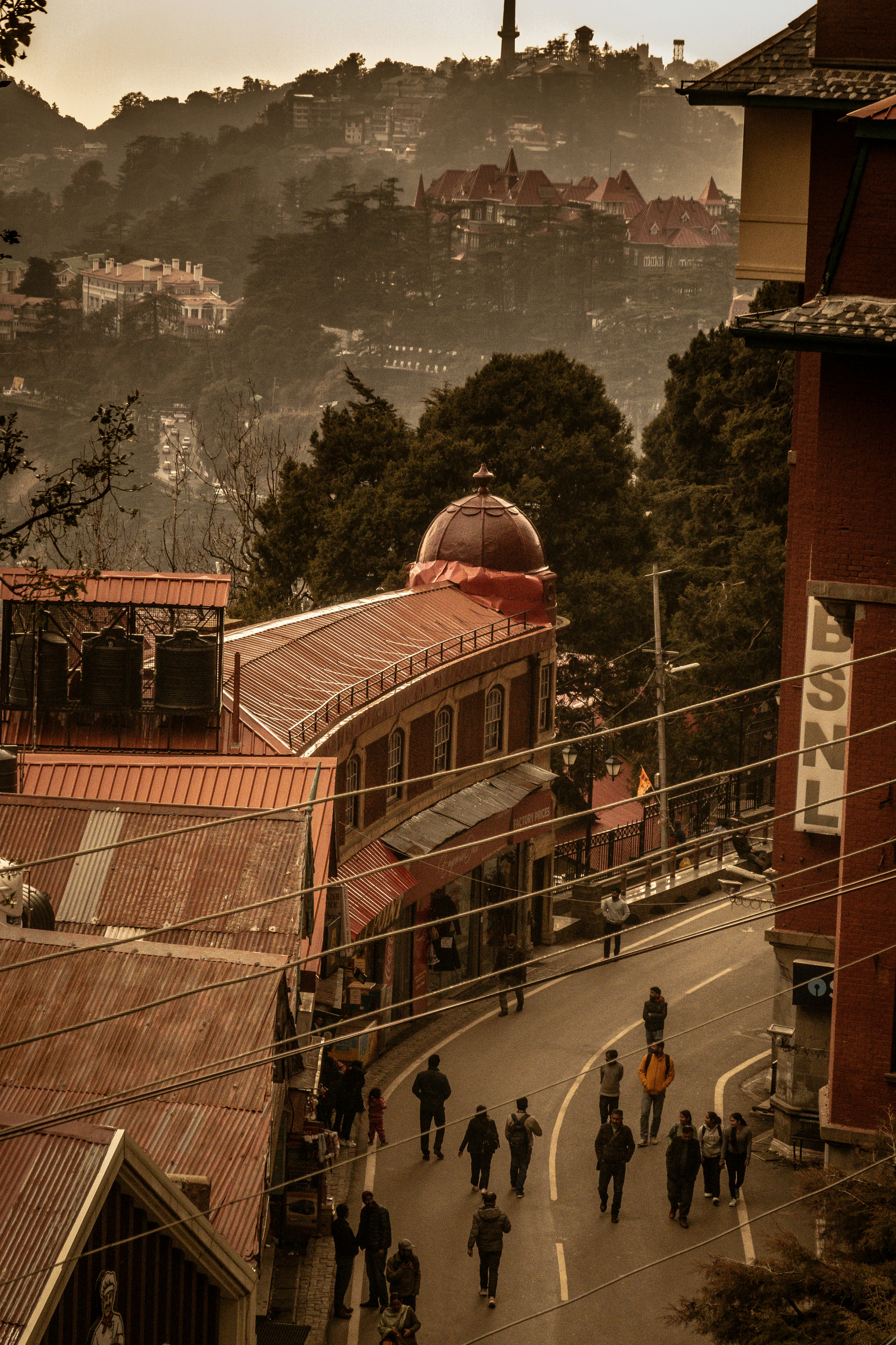 People walking on a winding street in a hilly town with historic architecture and lush greenery.