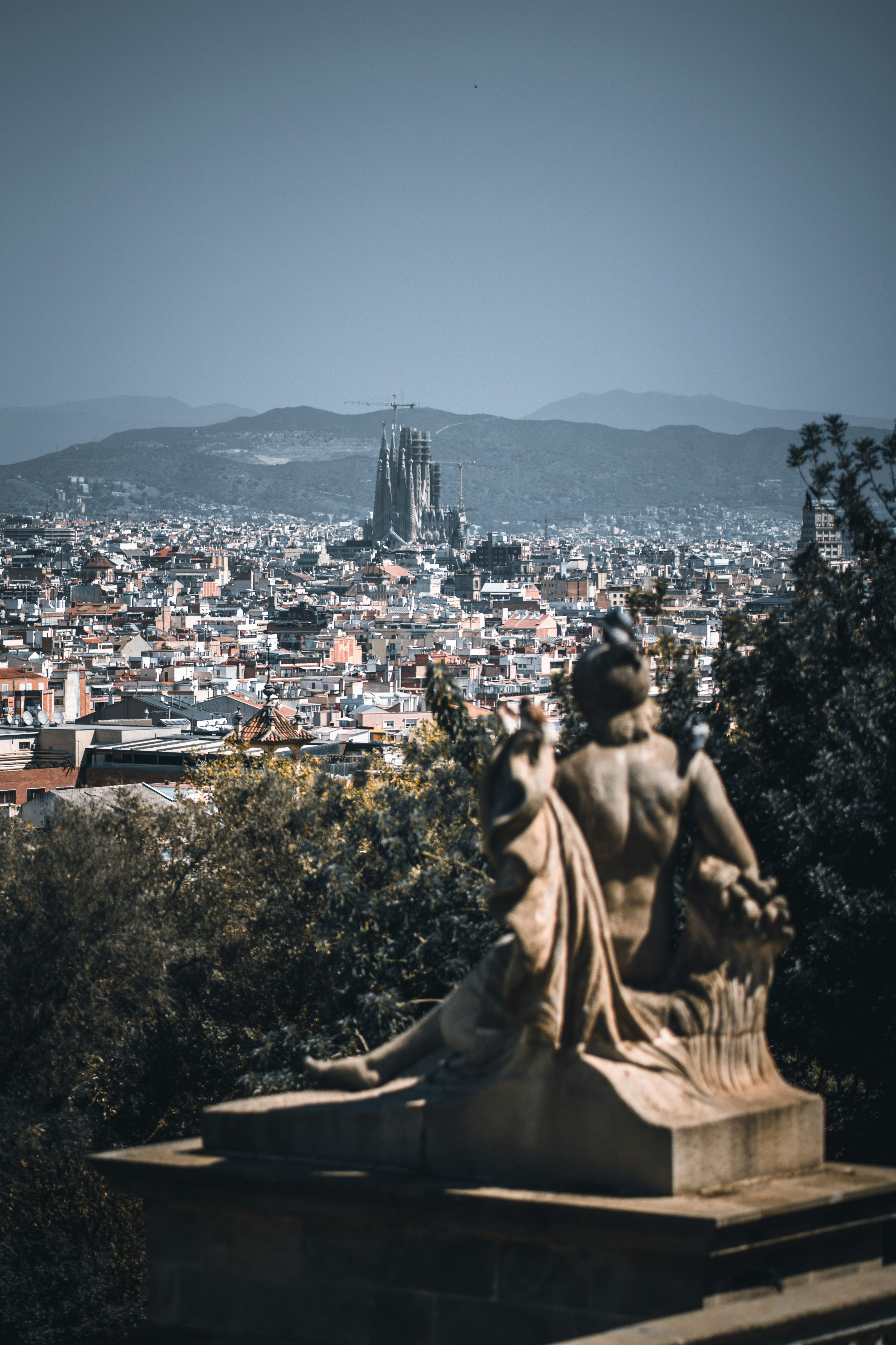 A stone statue gazes over the sprawling city of Barcelona, with the iconic Sagrada Família rising in the background amidst a hazy skyline.