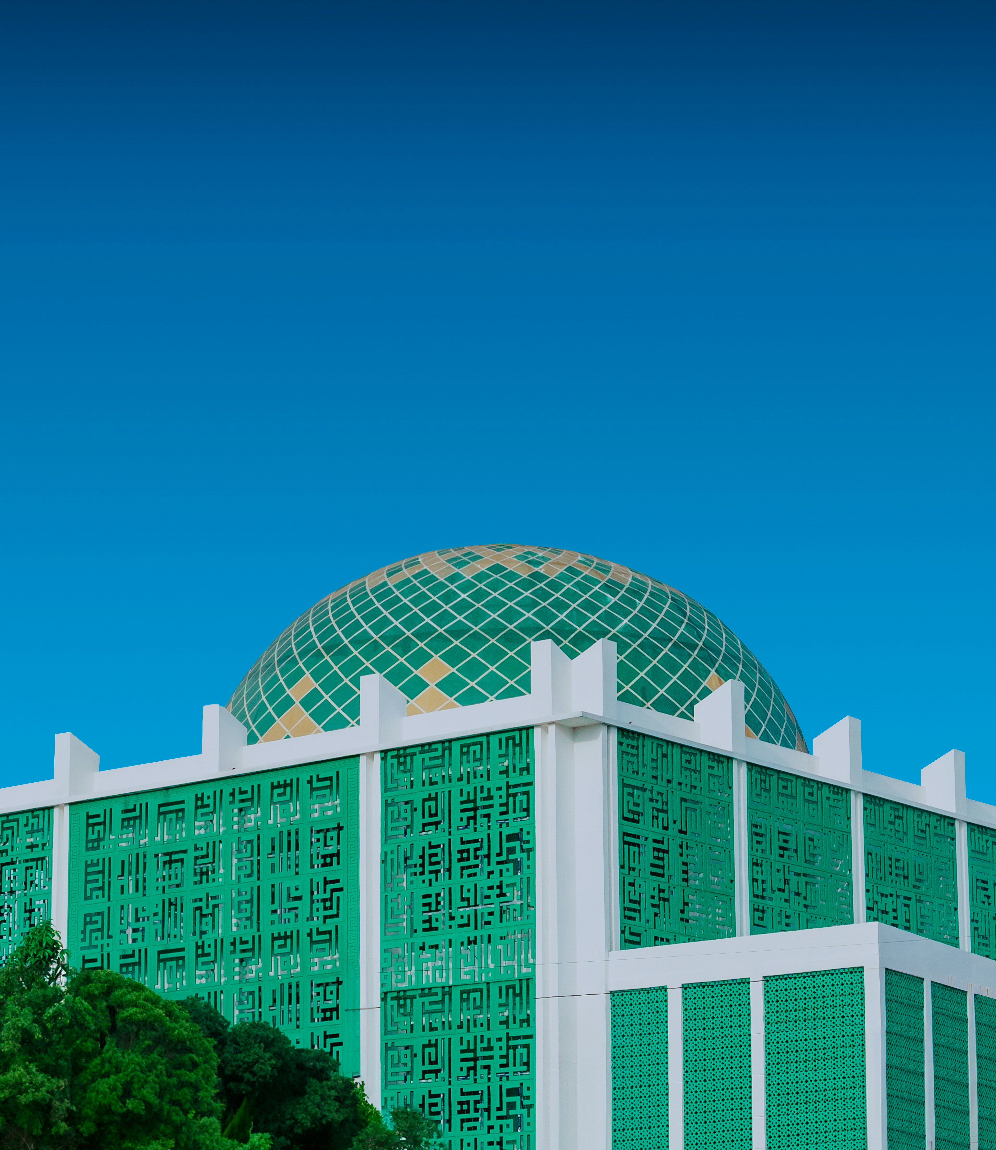 Architectural photograph of a teal-green building with patterned panels and a geodesic dome against a clear blue sky.