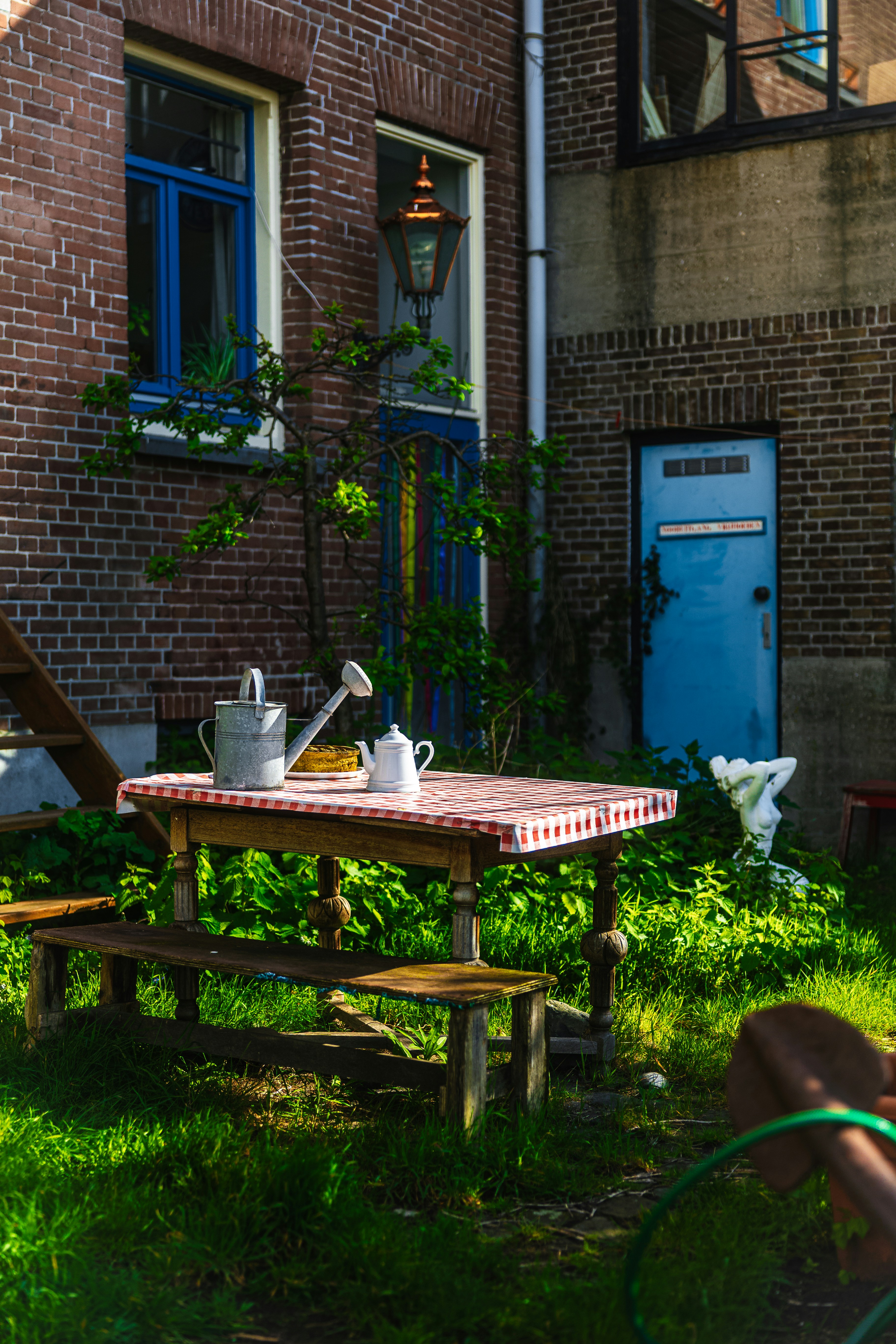 Teapot and Watering Cans on a Red-Checked Table in a Sunlit Brick Courtyard