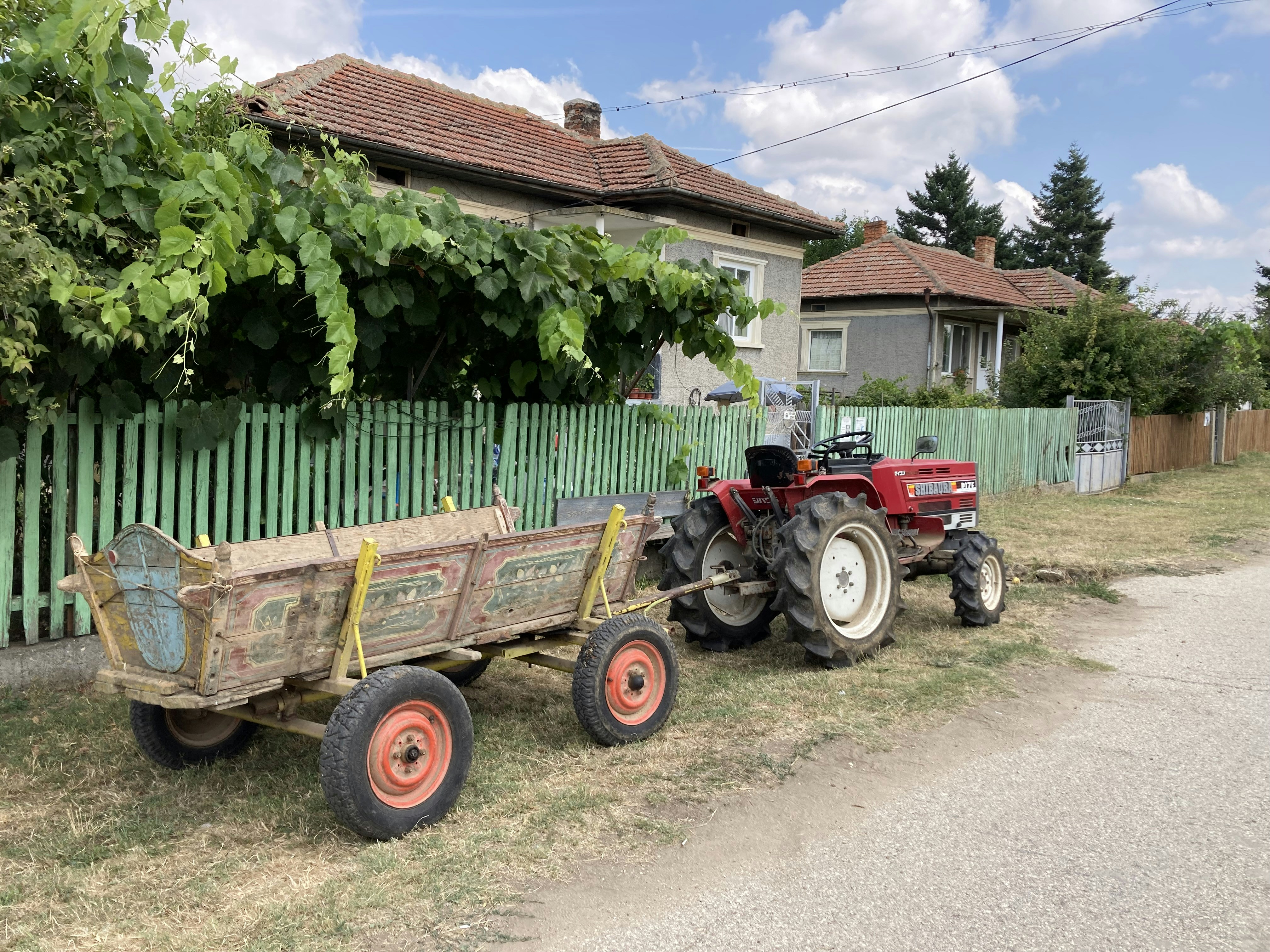 A vintage tractor parked next to a weathered wooden cart, framed by a lush green fence and a quaint house. This scene captures rural life in a charming setting.