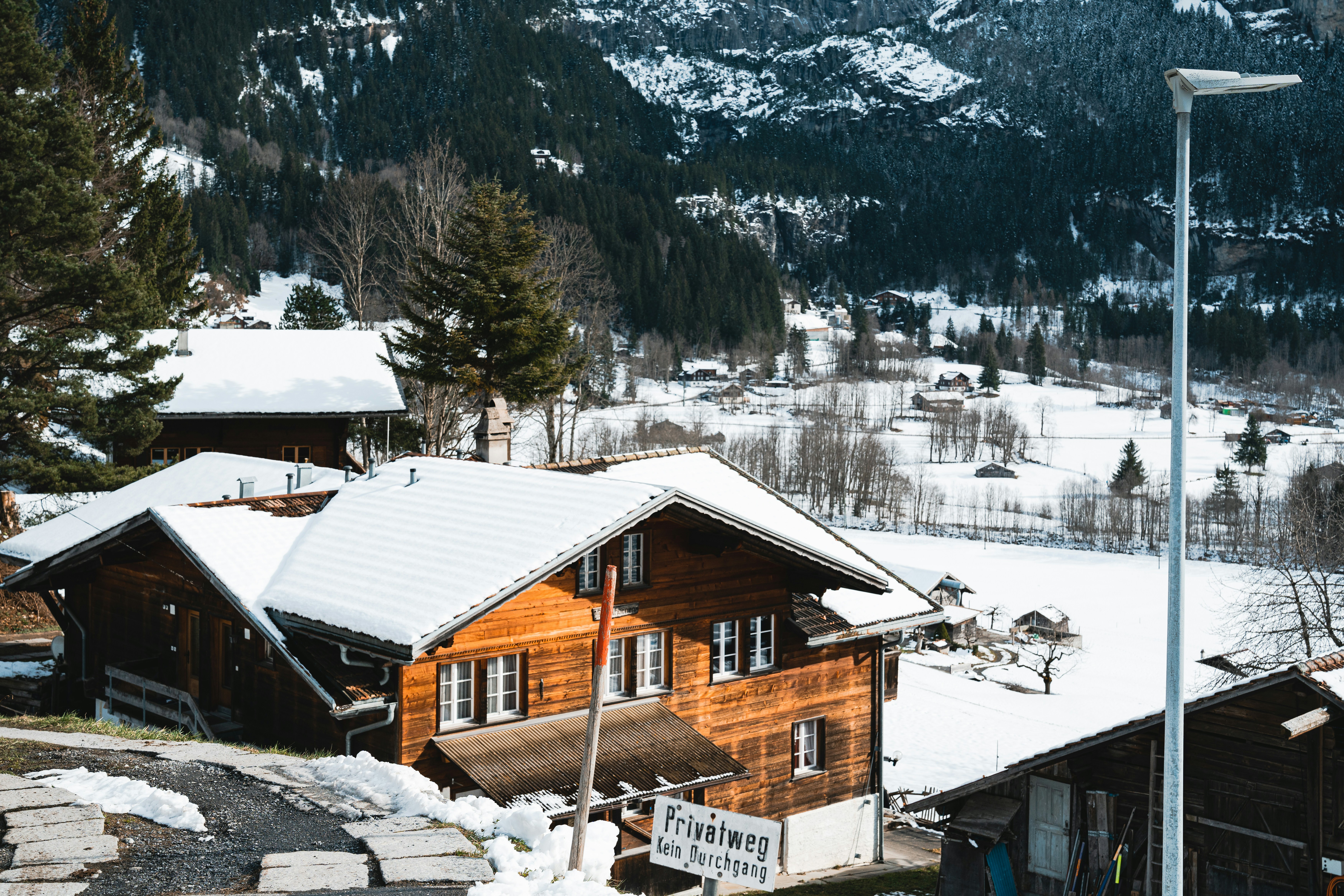 Traditional wooden chalet nestled in a snowy landscape, surrounded by evergreen trees and distant mountains. A signpost adds a touch of local charm.