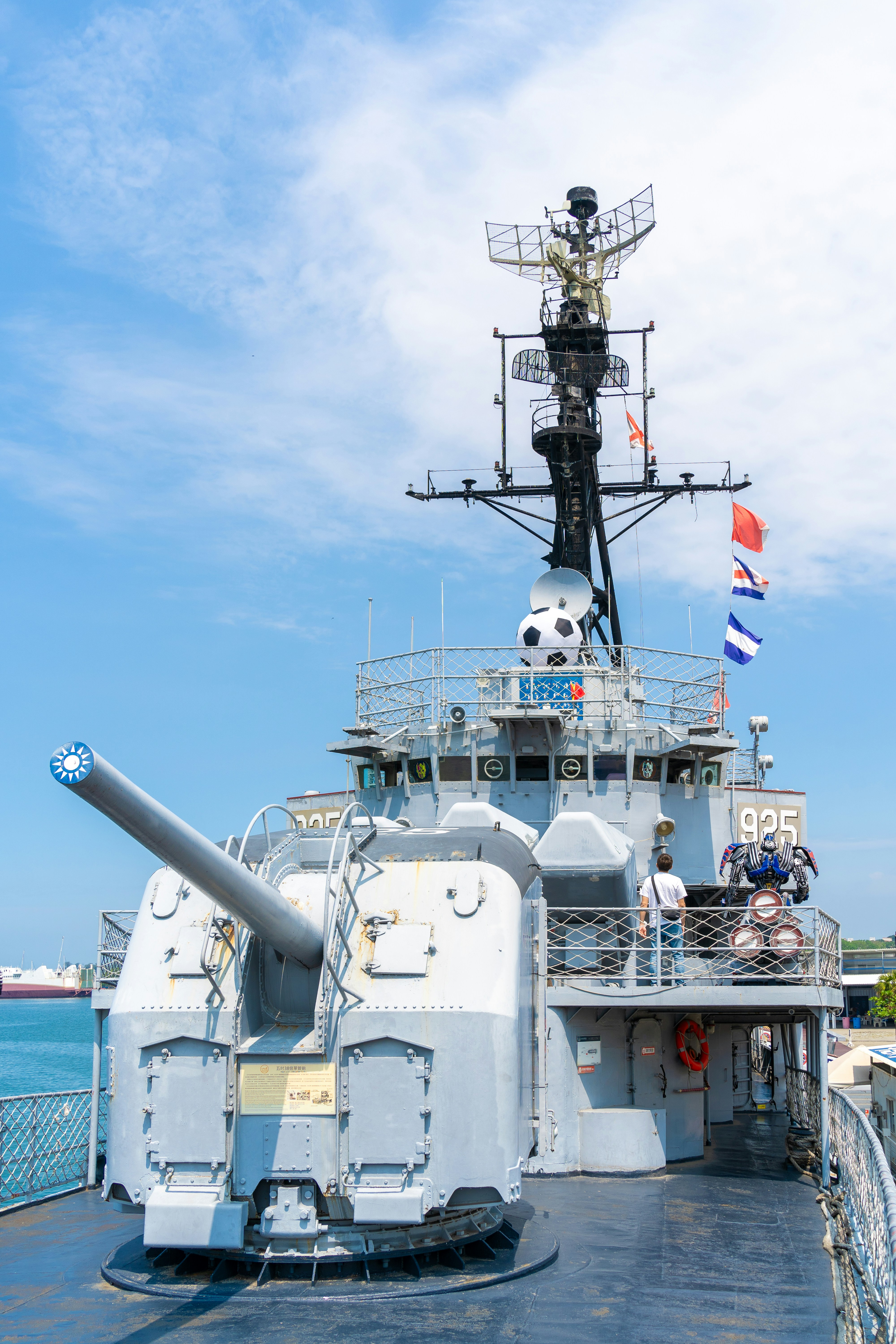 The deck of a naval ship showcases its formidable cannon and radar, with crew members engaged in activity against a backdrop of blue skies and distant vessels.
