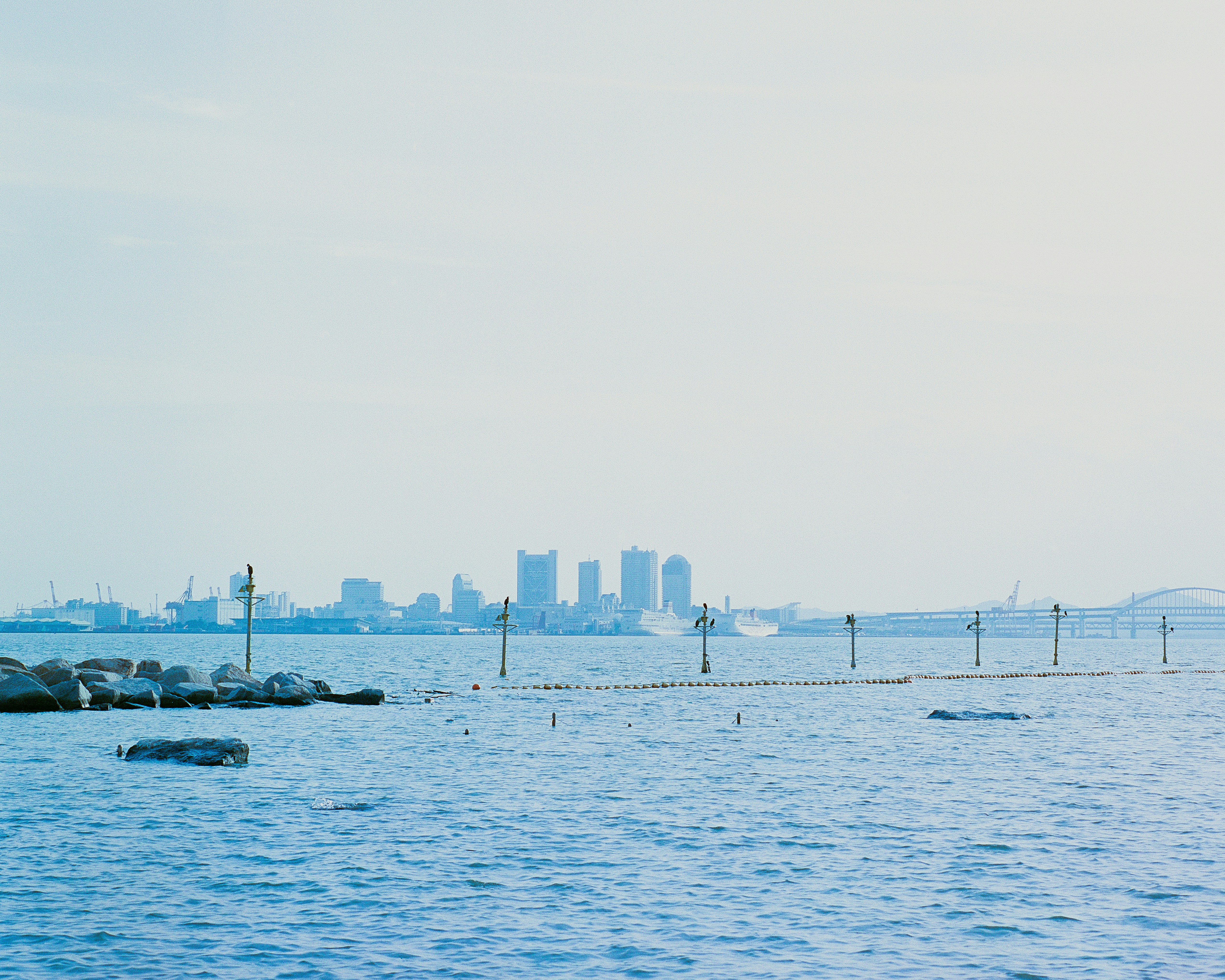Distant city skyline viewed over calm ocean with scattered rocks and buoys.