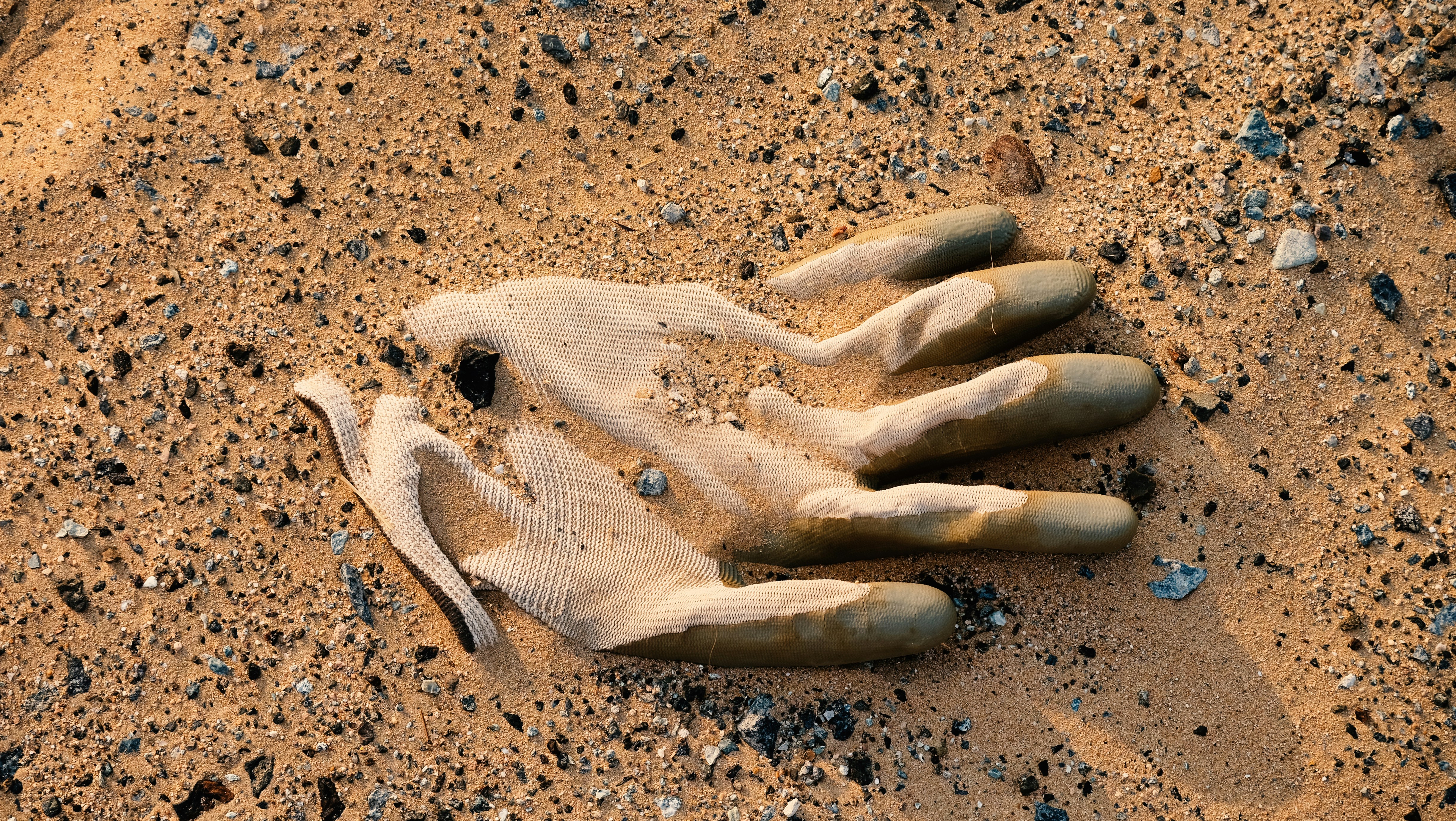 A weathered glove rests on a sandy surface, surrounded by small stones and debris, highlighting the intersection of human presence and nature's reclaiming force.