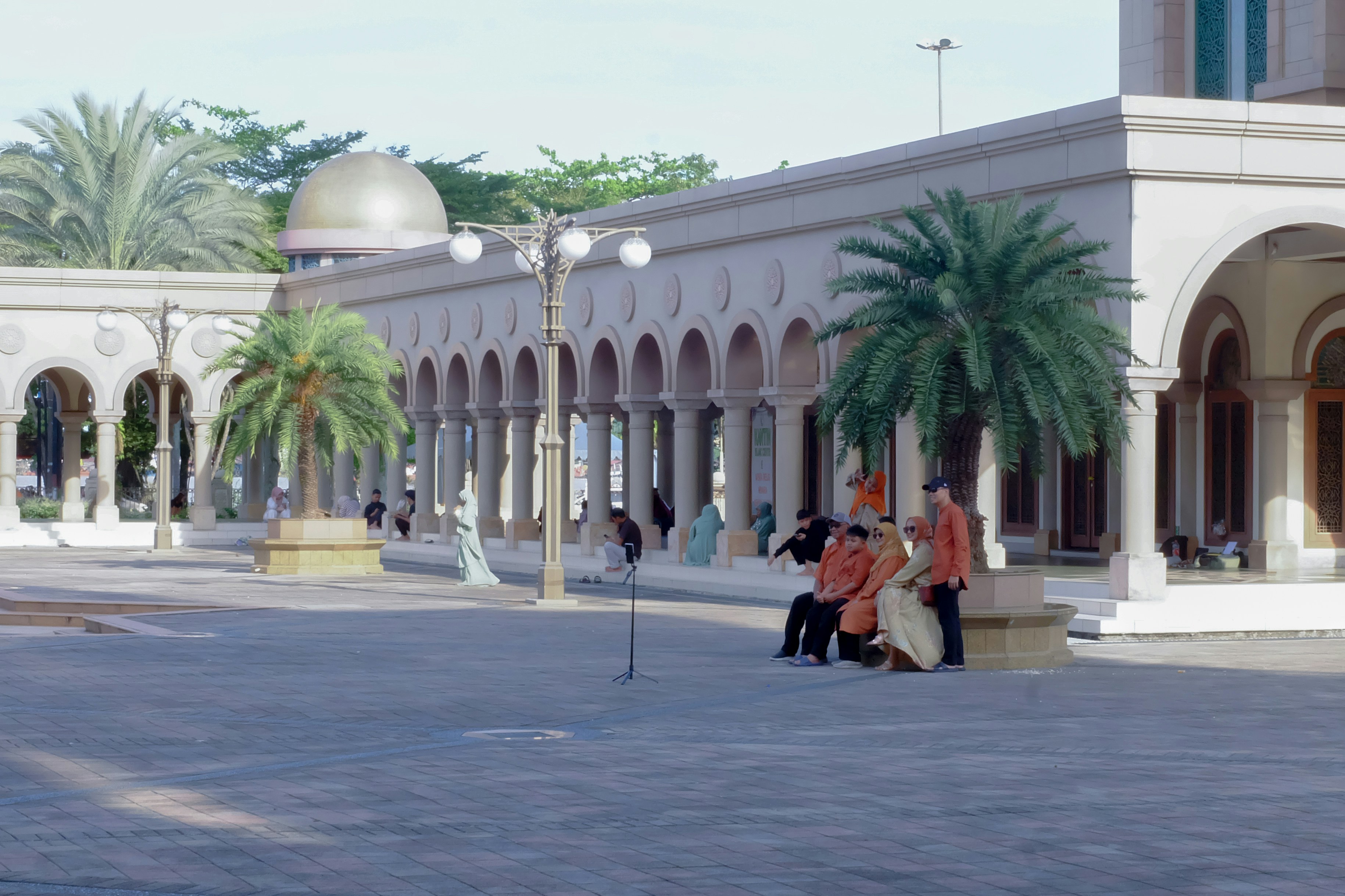 People gather near palm trees and arched colonnades in a sunlit plaza.