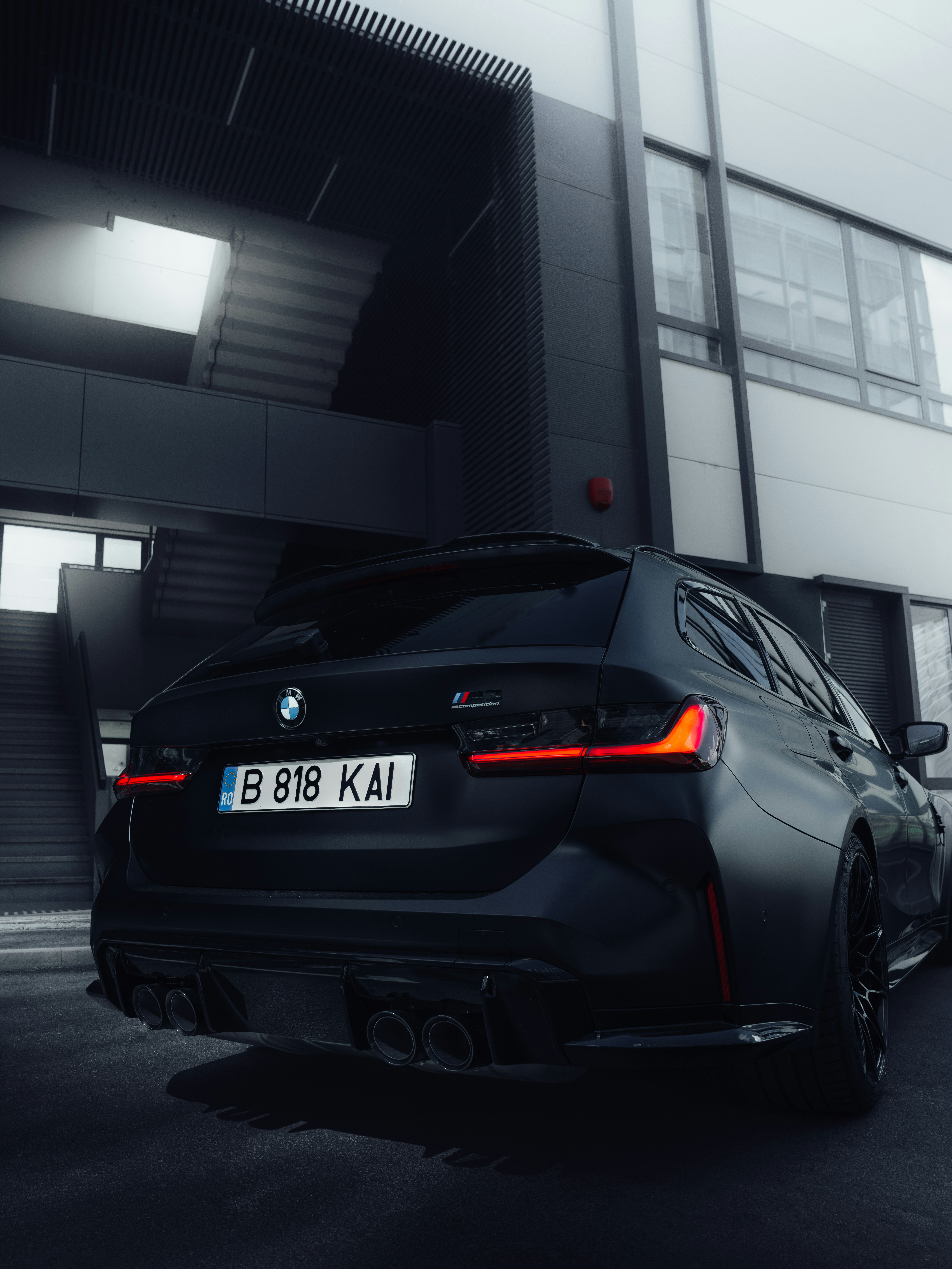 Dark BMW M rear view parked beside a modern industrial building, emphasizing red taillights and quad exhausts. Moody lighting and architectural lines frame the scene.