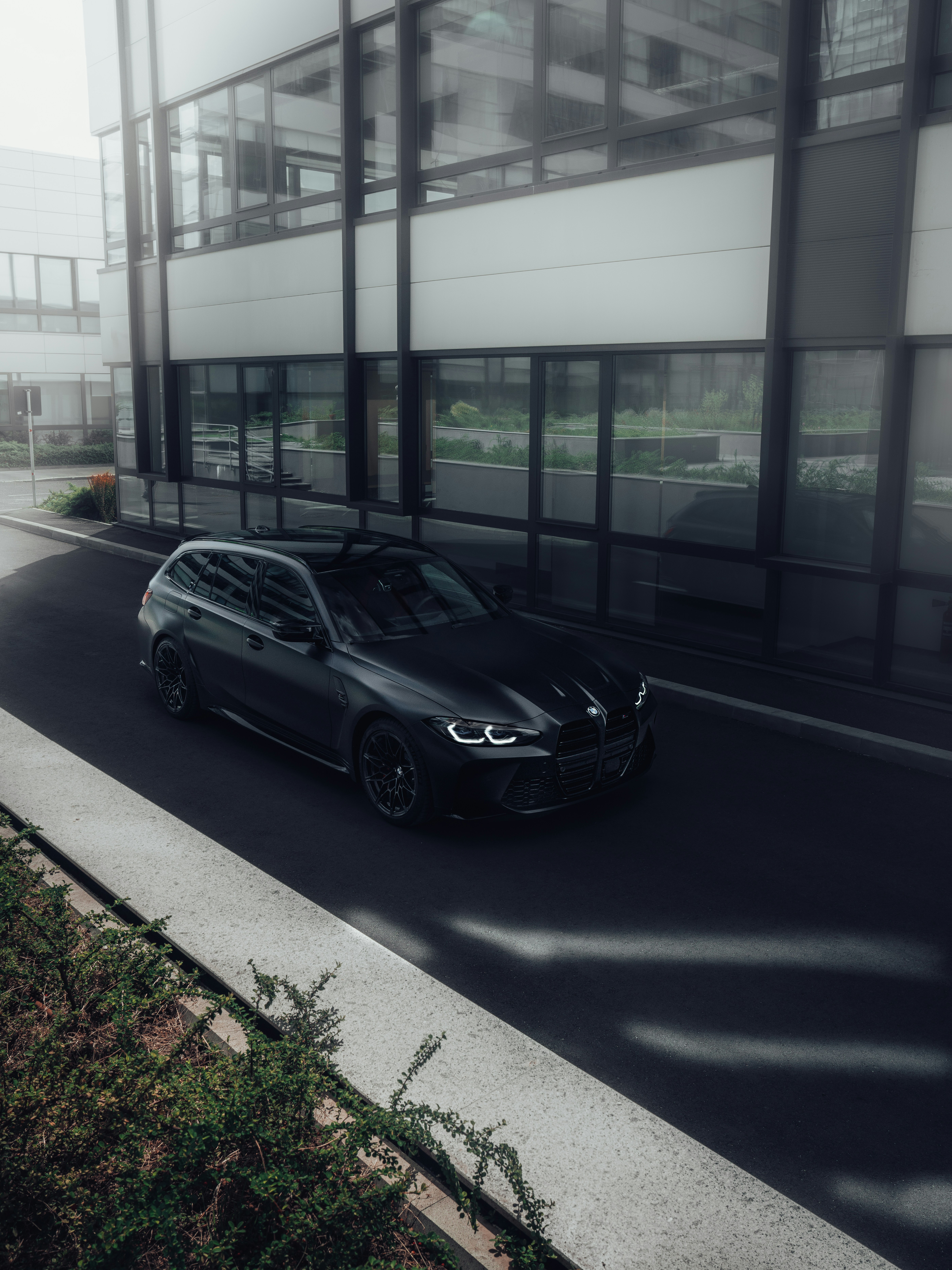 Photograph shows a jet-black BMW sedan parked along a sunlit urban street beside a glass-walled office building.