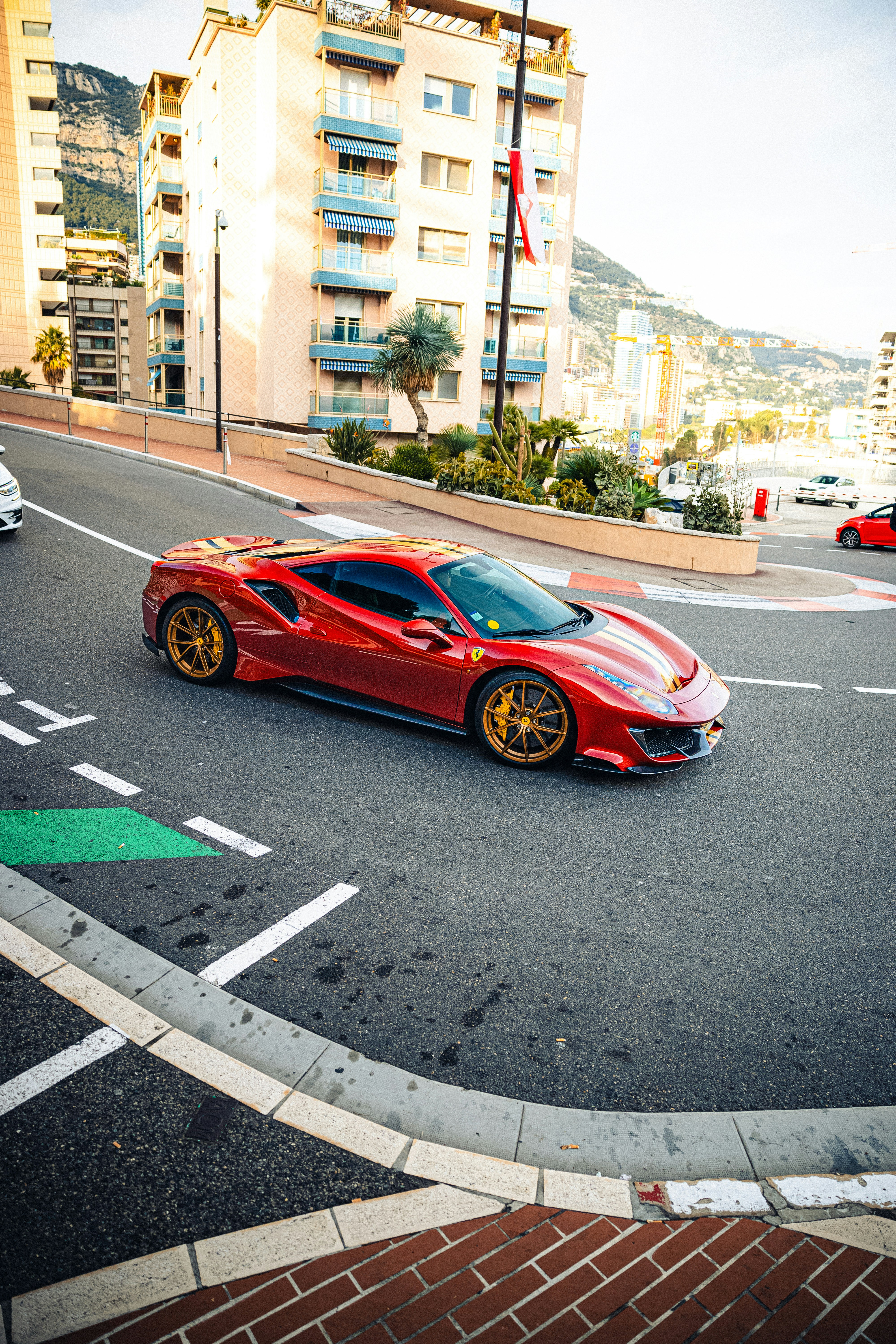 Sleek crimson Ferrari turns through a sunlit bend, framed by pastel apartment blocks and a distant hillside.