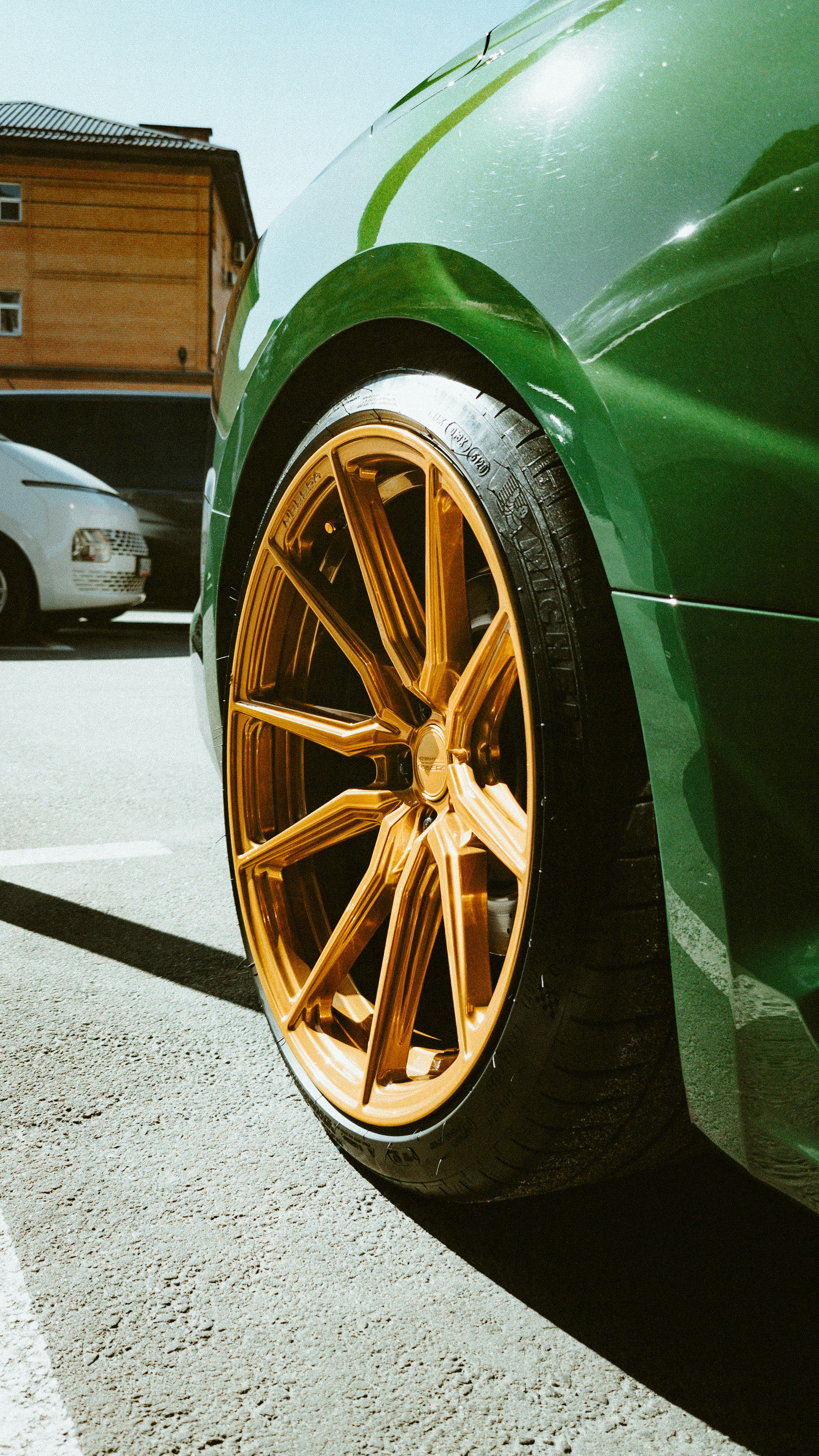 Close-up car photograph focusing on a glossy green body and a gold alloy wheel, captured from a low angle. The composition highlights wheel design and reflective surfaces against a daylight backdrop.