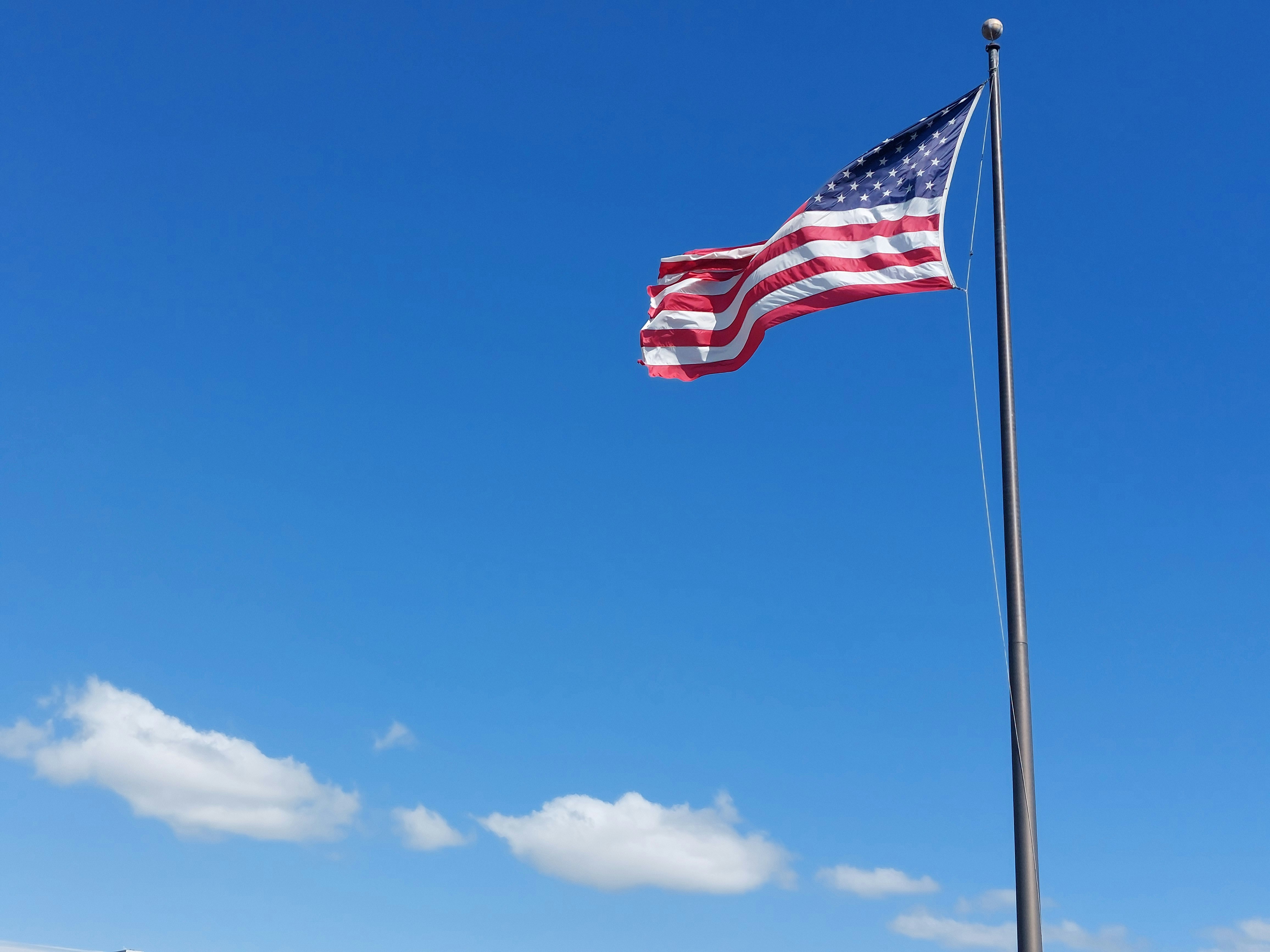 American flag on a tall flagpole waves against a bright blue sky with scattered clouds.