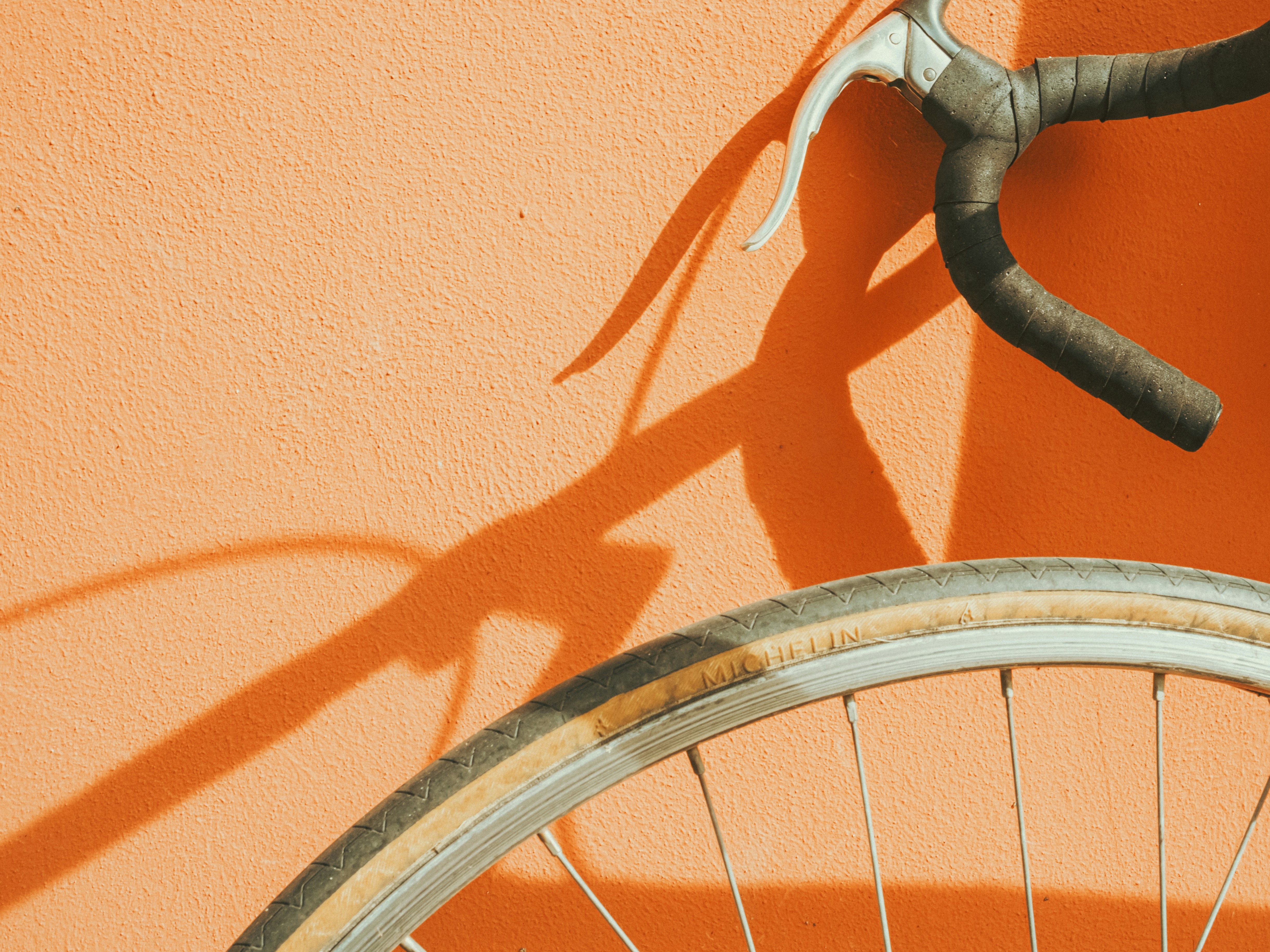 Photograph showing a bicycle wheel and handlebars casting bold shadows on a warm orange wall. The composition emphasizes geometric lines and color contrast.