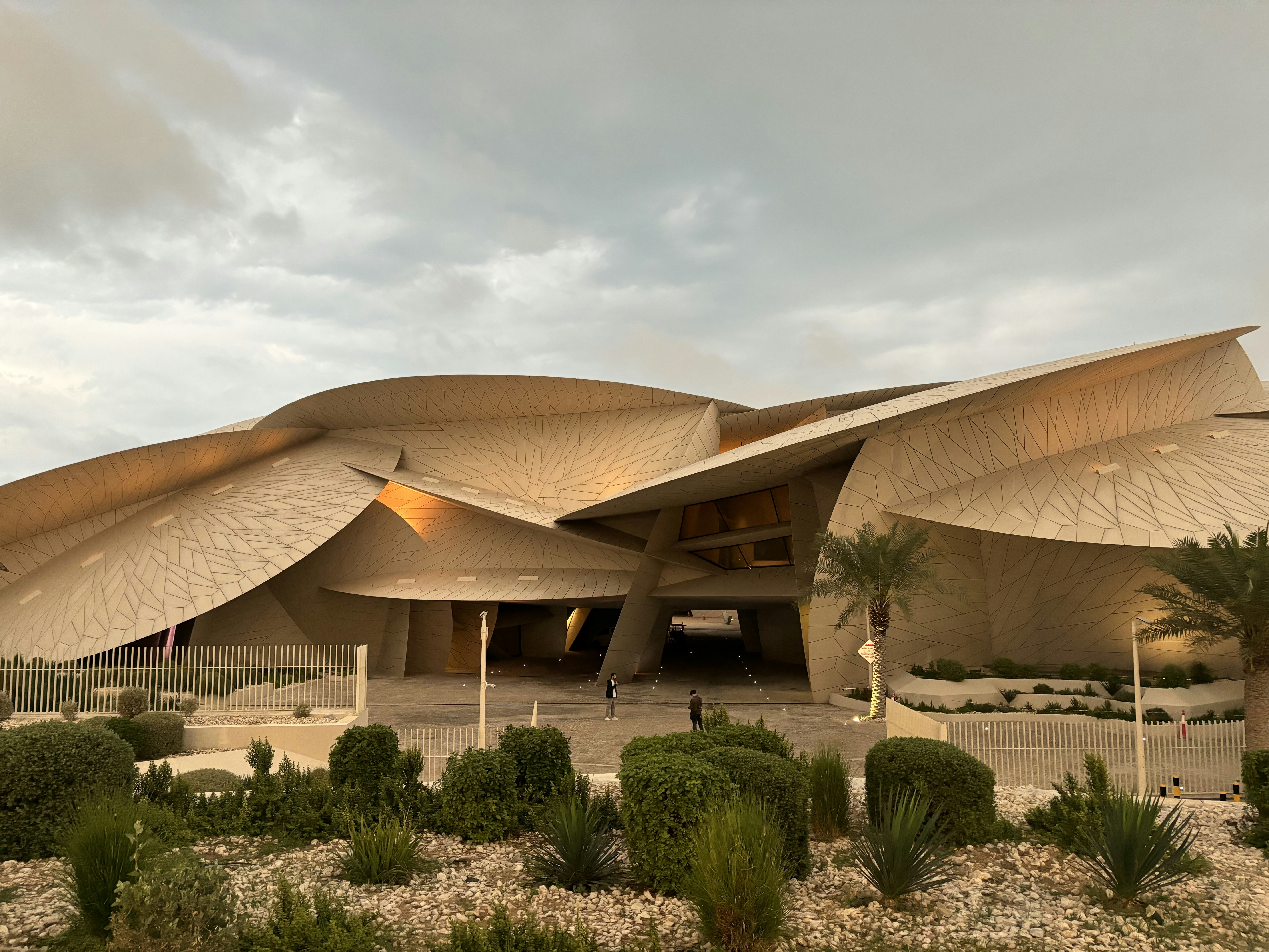 Photograph of a sculptural desert plaza featuring sweeping concrete canopies and angular supports. Palm-lined forecourt and pebble landscaping lead the eye to a shaded entrance.