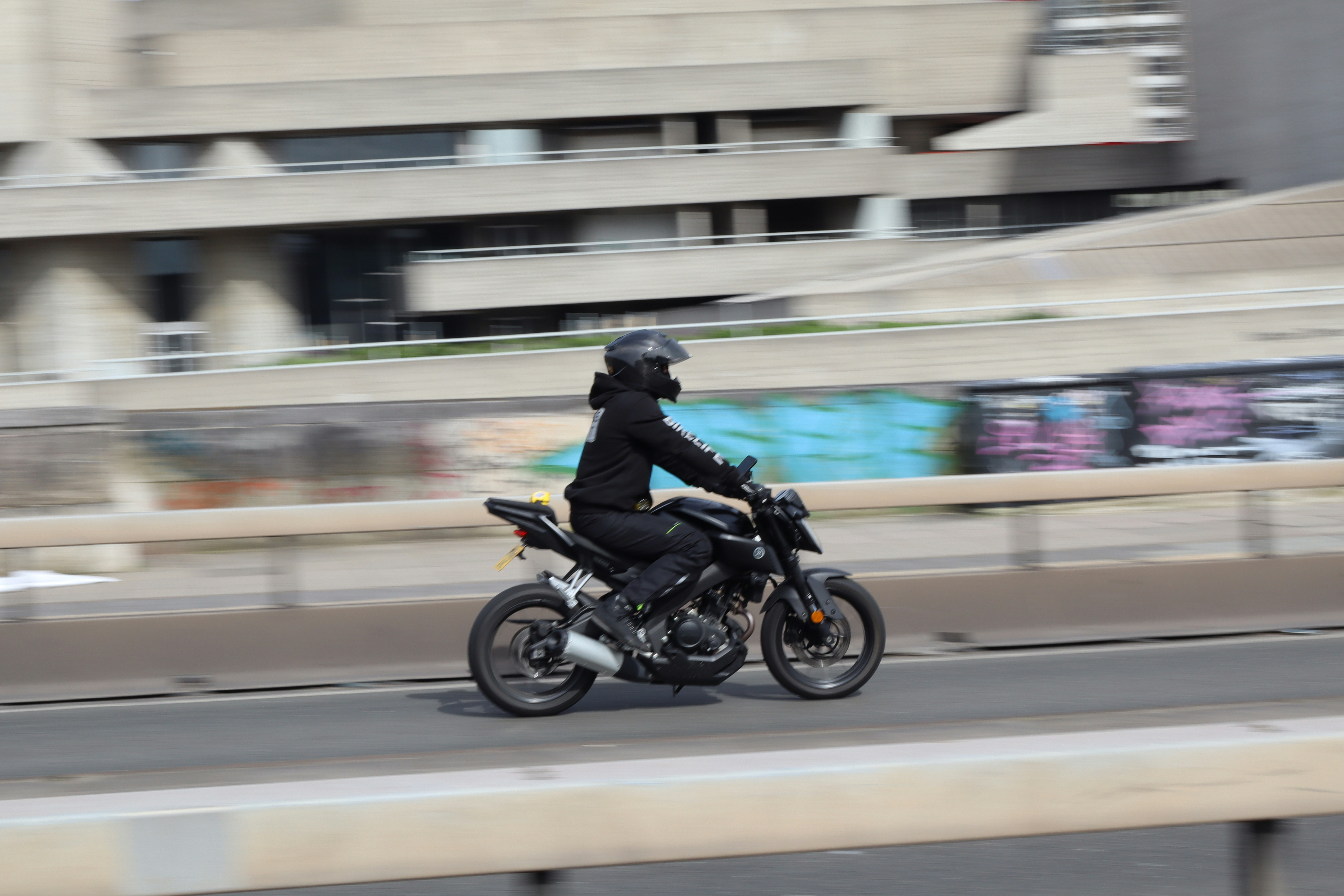 Motorcyclist in black gear speeding along a city roadway with blurred background.