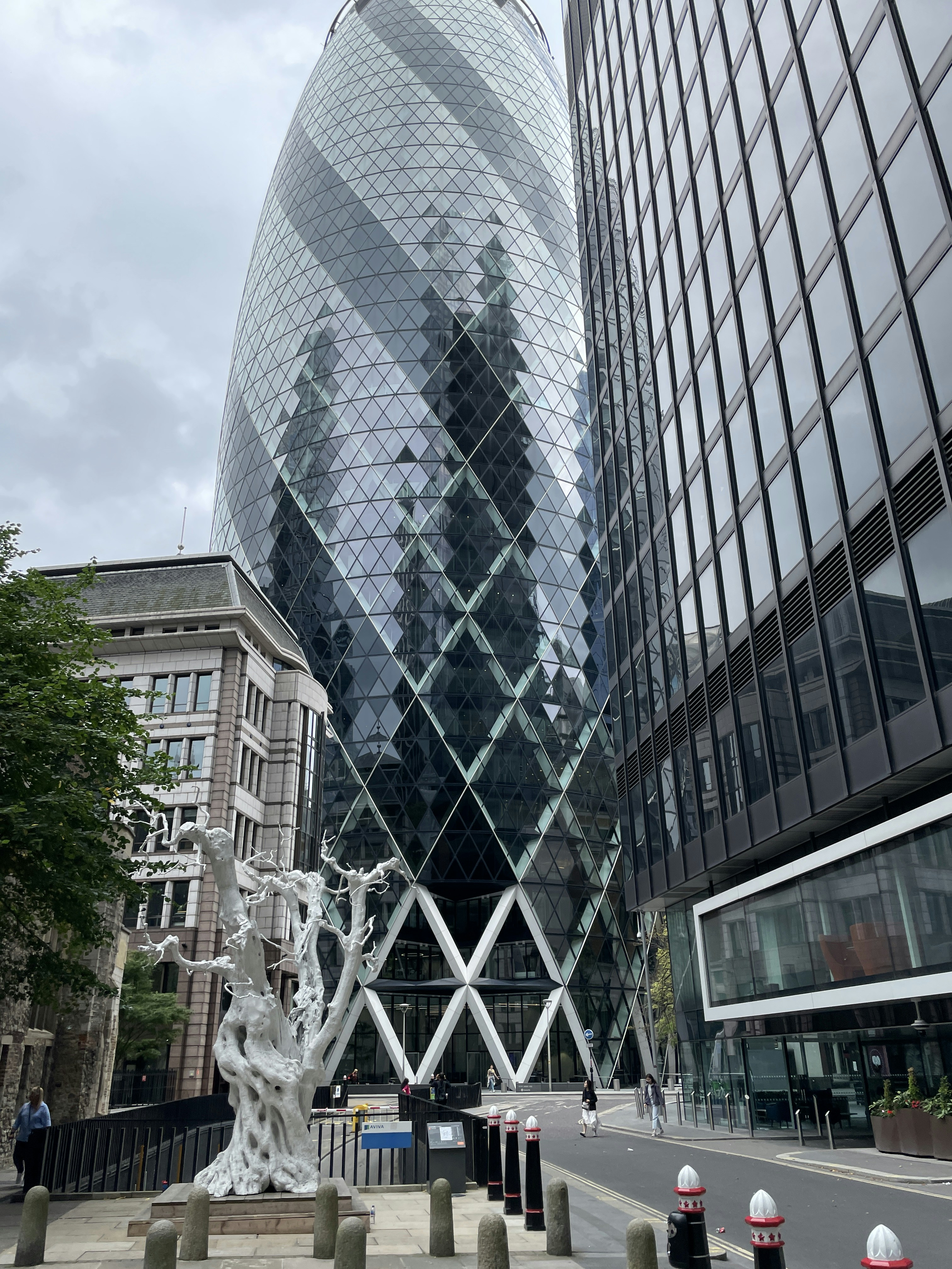 A striking modern skyscraper reflects the surrounding architecture and trees, showcasing the blend of nature and urban design. A white sculptural tree stands prominently in the foreground.