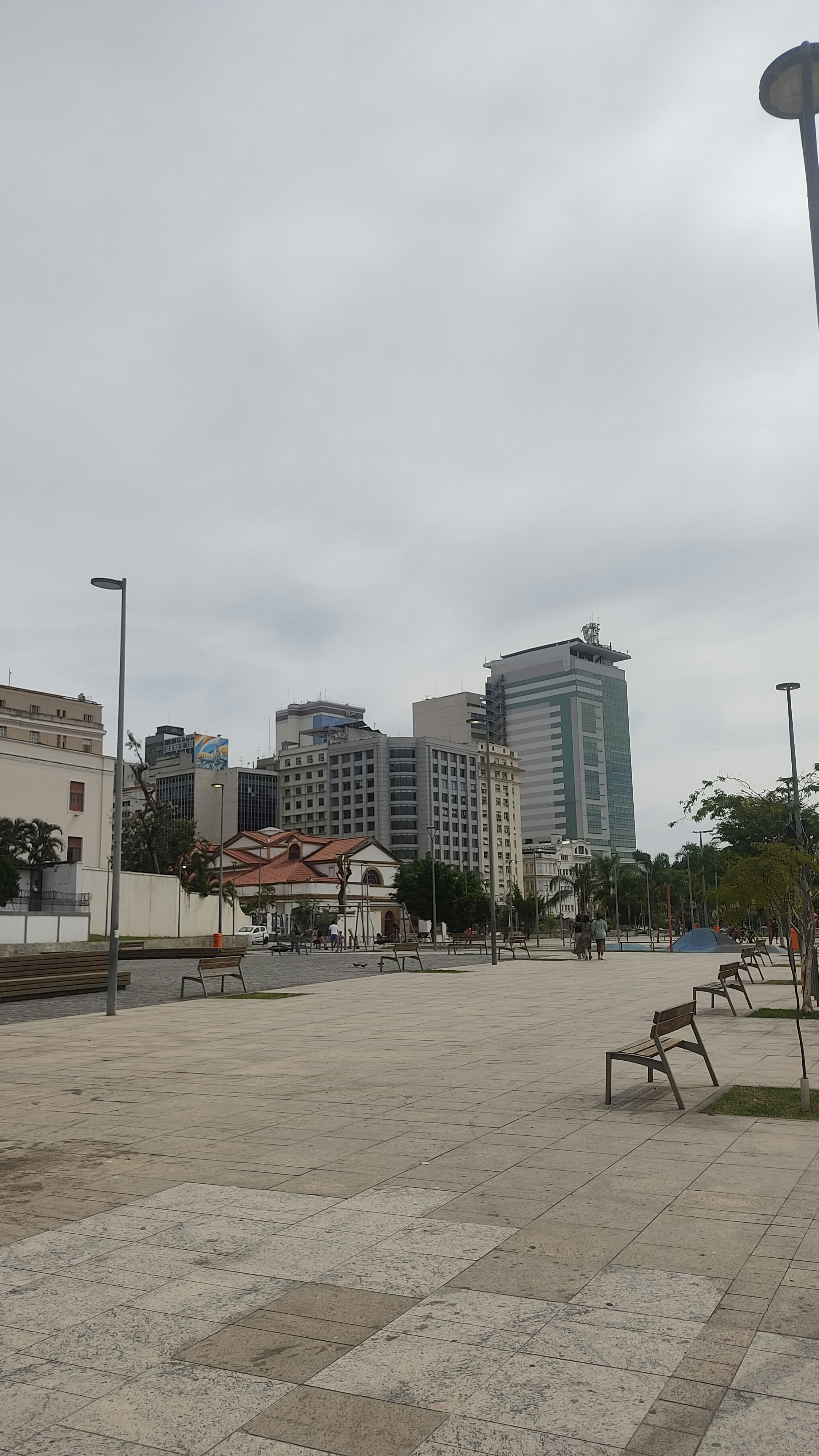 Modern cityscape with benches in a spacious park, framed by towering buildings under a cloudy sky.