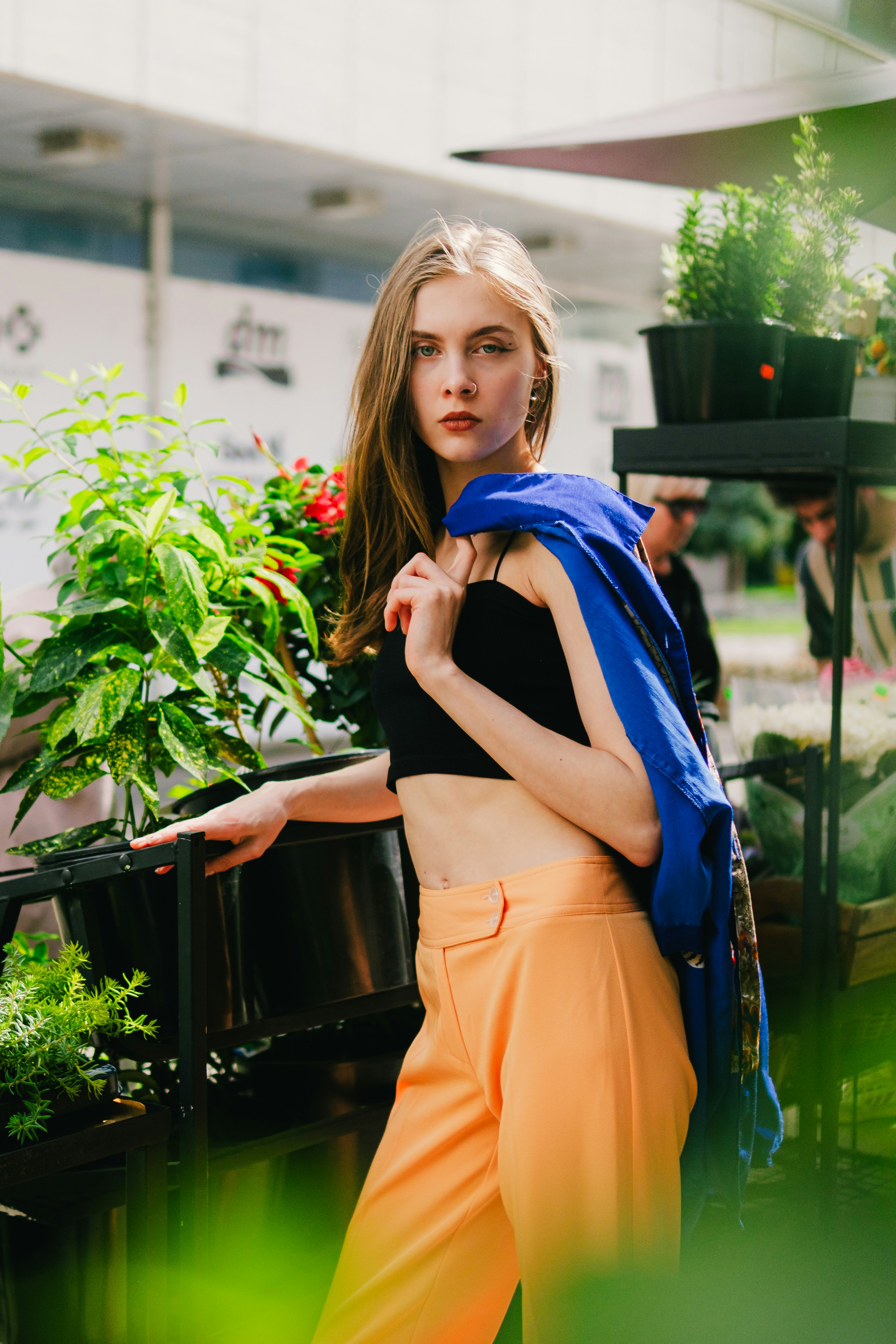 Fashion portrait of a young woman in a plant-filled greenhouse setting. She wears a black crop top, peach-orange pants, and has a blue jacket draped over her shoulder.