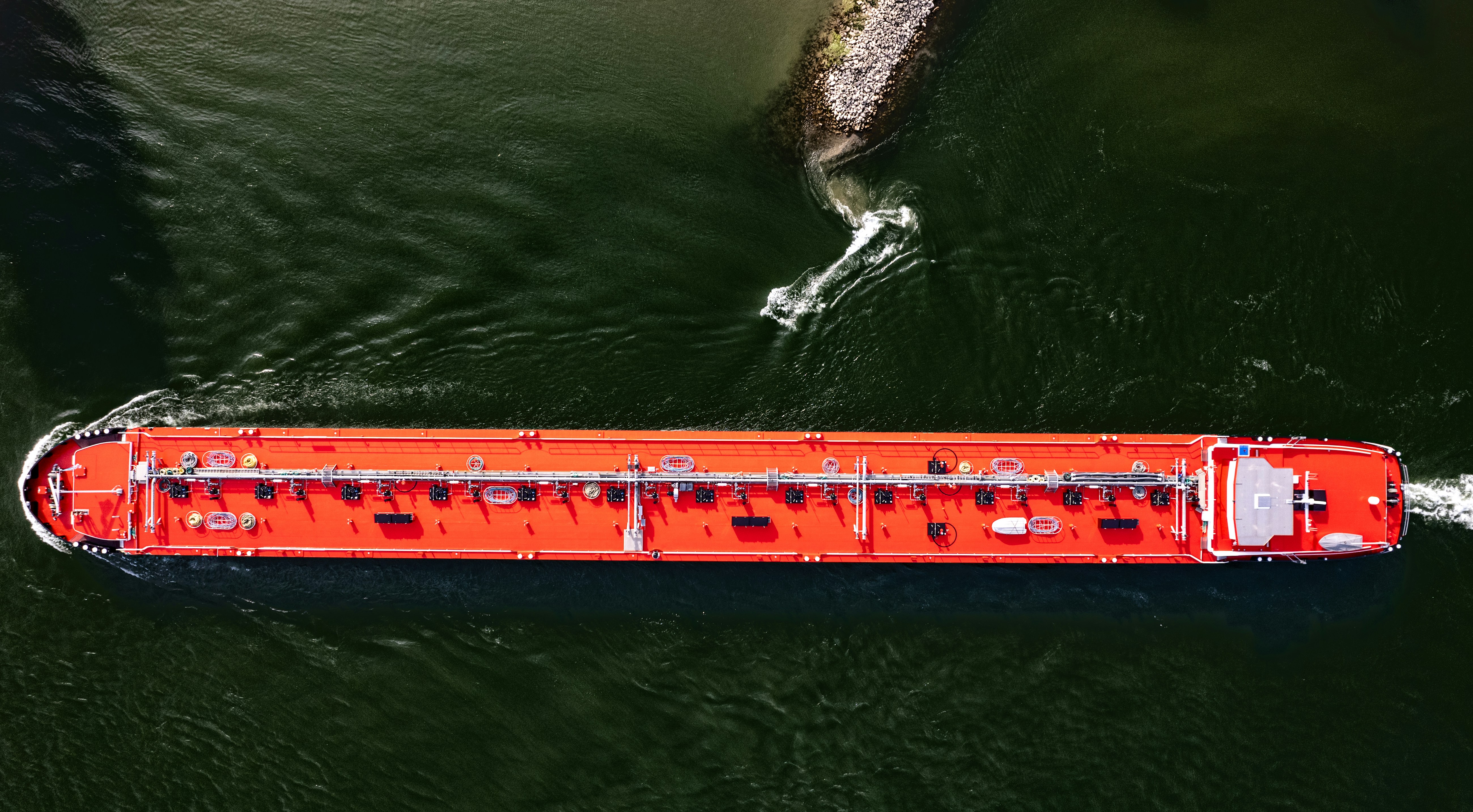 An aerial shot captures a striking red cargo ship navigating through a waterway, its path evident by the wake in the green water, showcasing the vessel's role in the complex system of maritime logistics and trade.