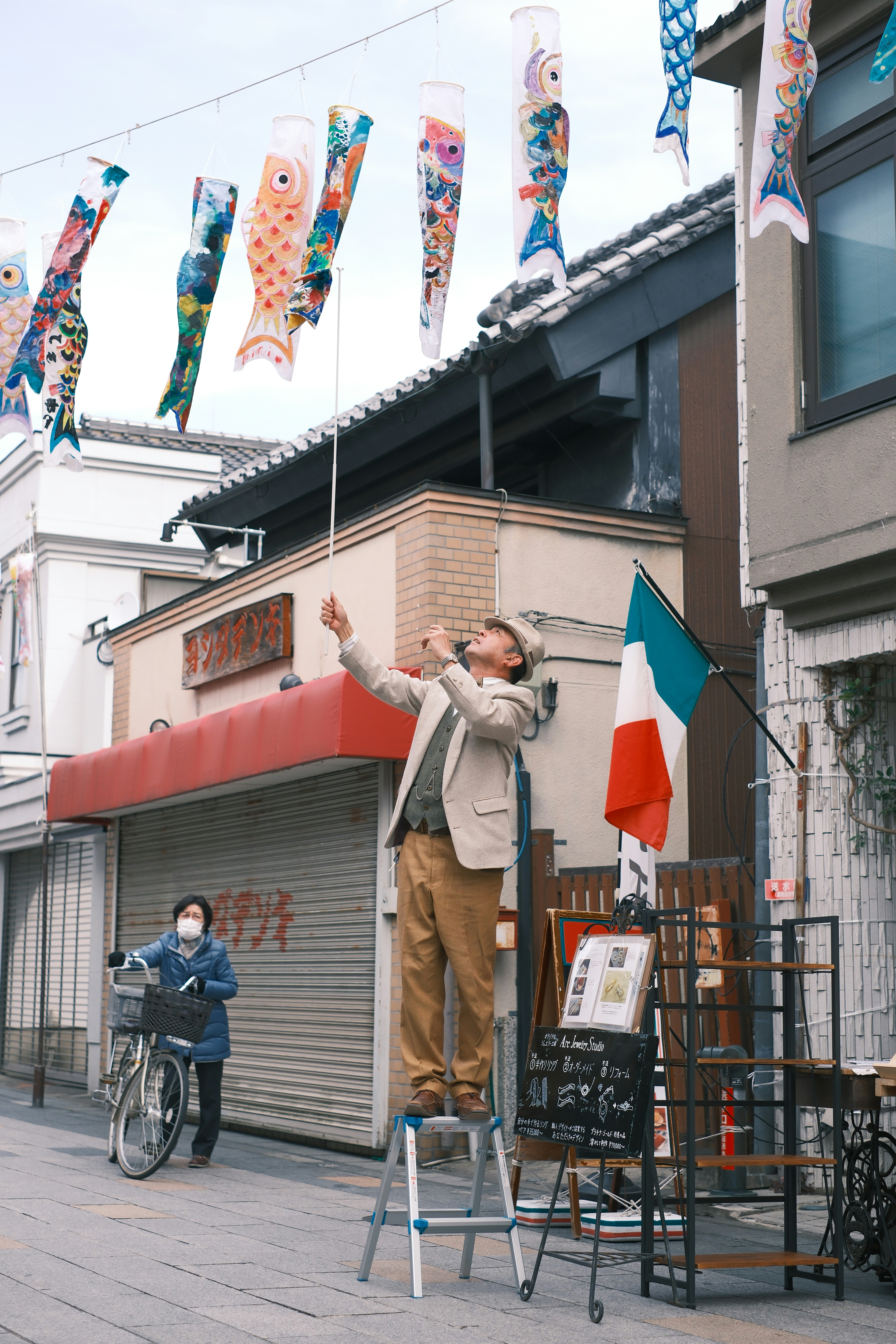 Street-side acrobat in a festival of koi winds: man on a step stool reaches for airborne banners