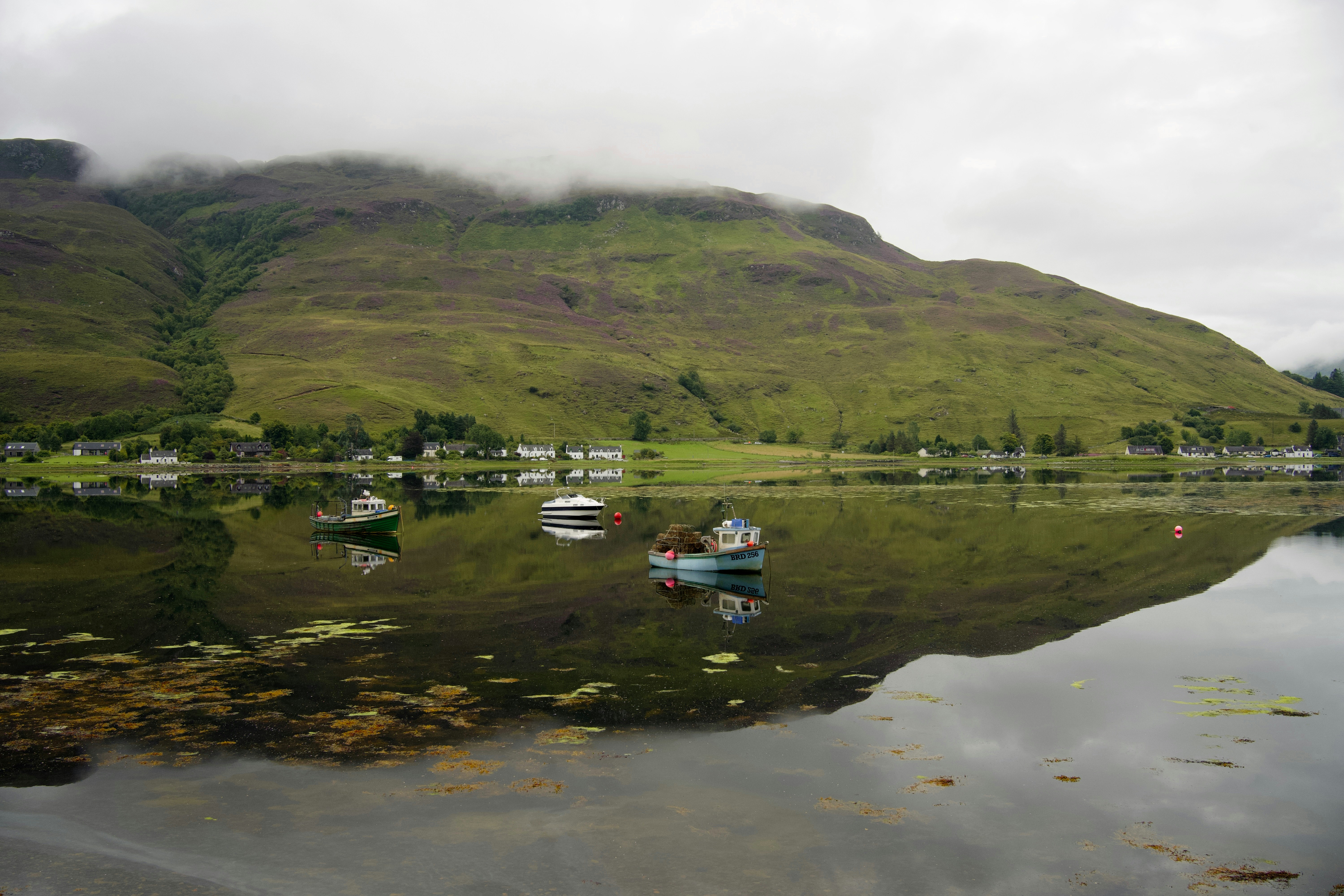 Boats float on a tranquil loch with mirror-like reflections beneath overcast skies and lush hills in Scotland.