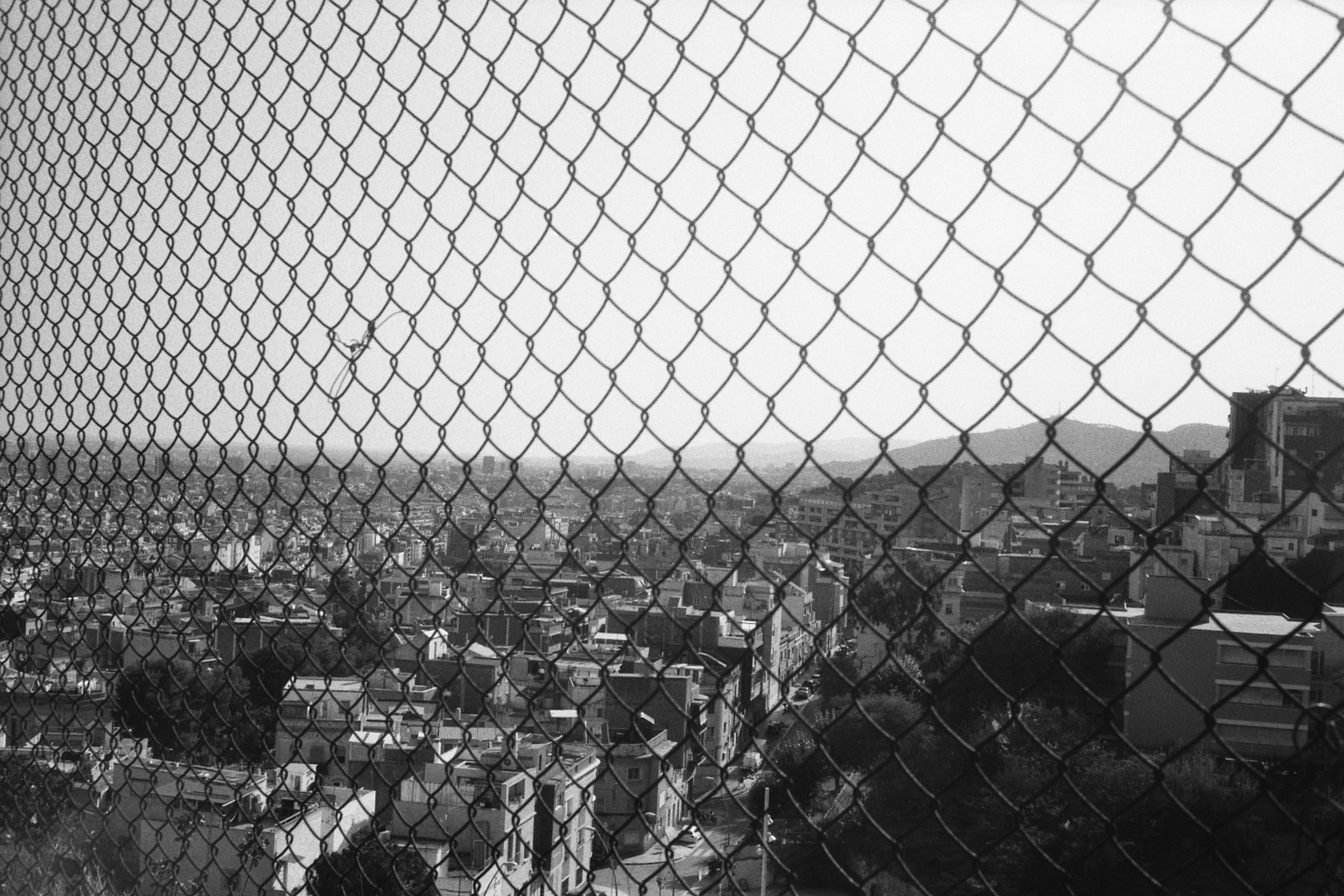 Cityscape framed by a chain link fence with distant hills on the horizon.