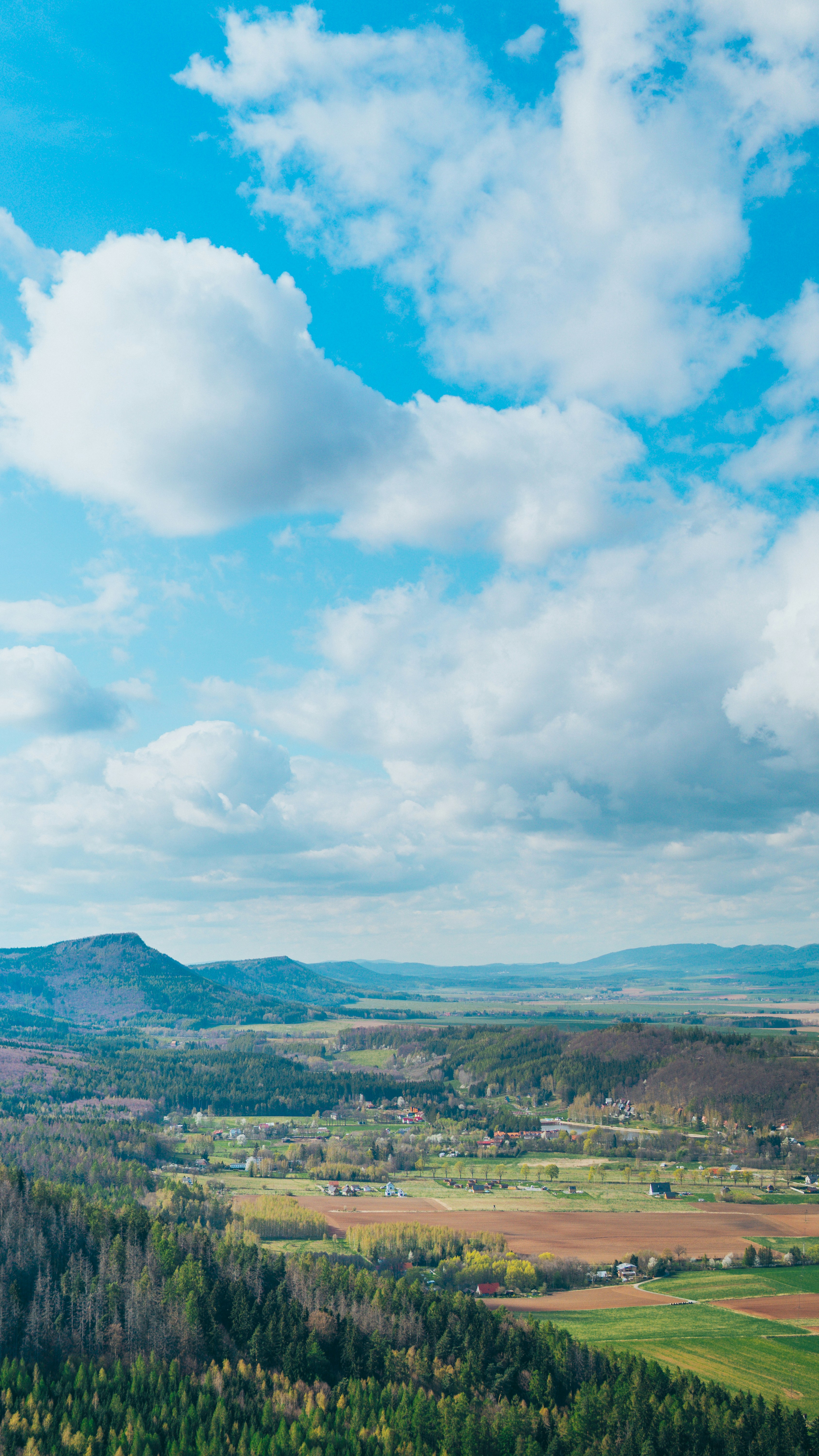 Expansive landscape featuring lush fields and rolling hills, framed by a vibrant blue sky dotted with fluffy clouds.