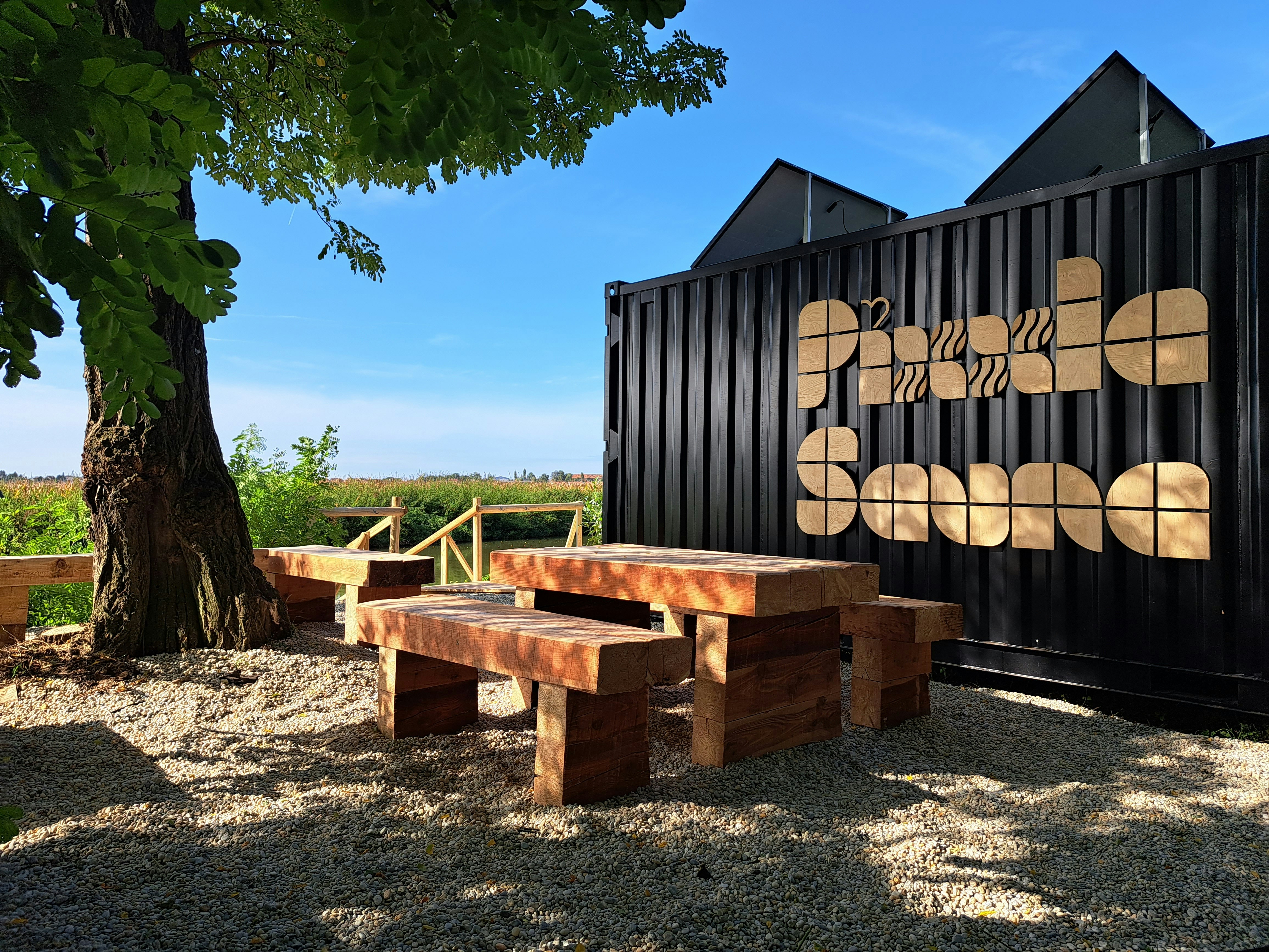 Sunlit outdoor seating area with chunky wooden benches beside a black wall bearing large beige block letters.