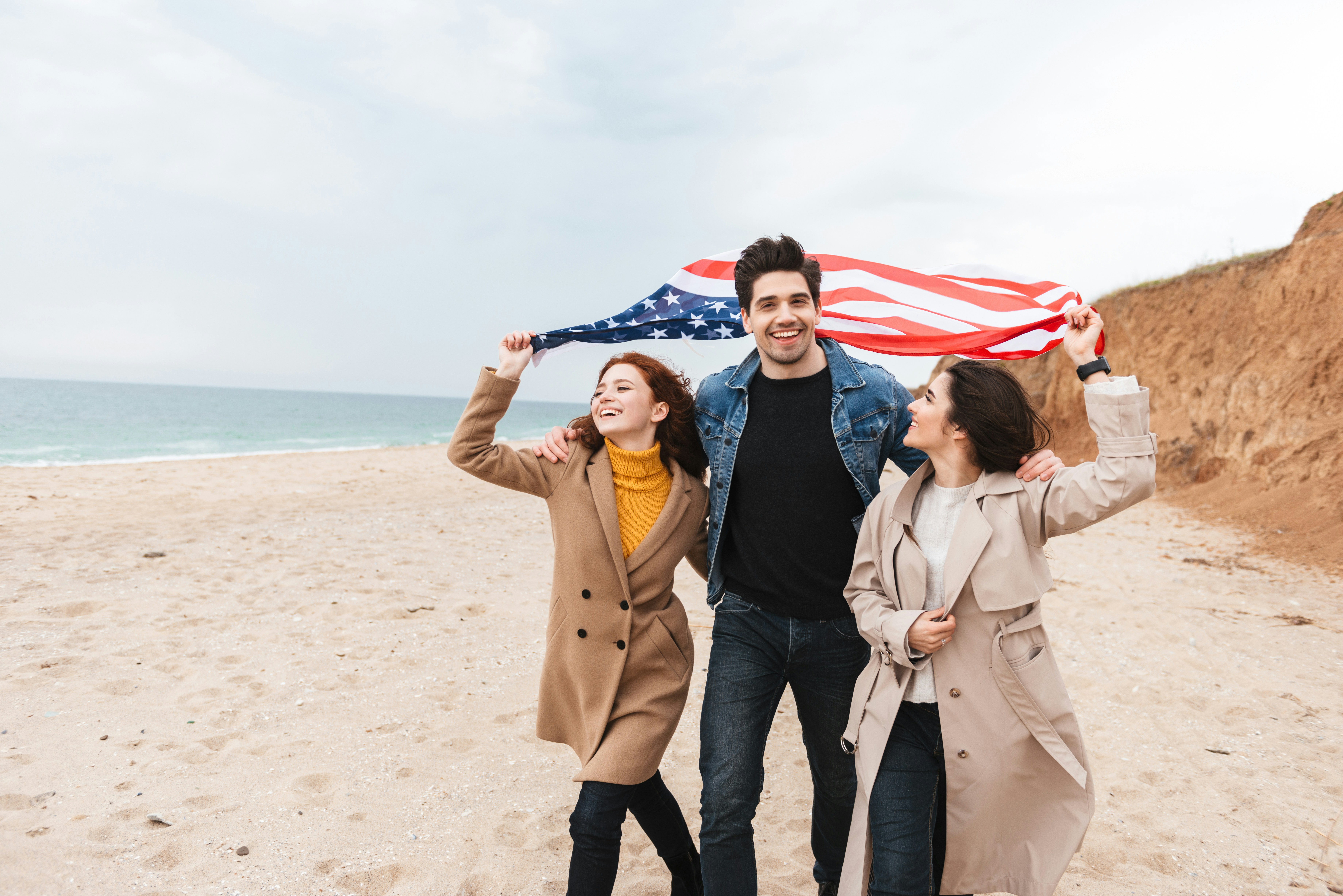 Coastal Cheer: Three Friends Rally with the American Flag on a Quiet Beach