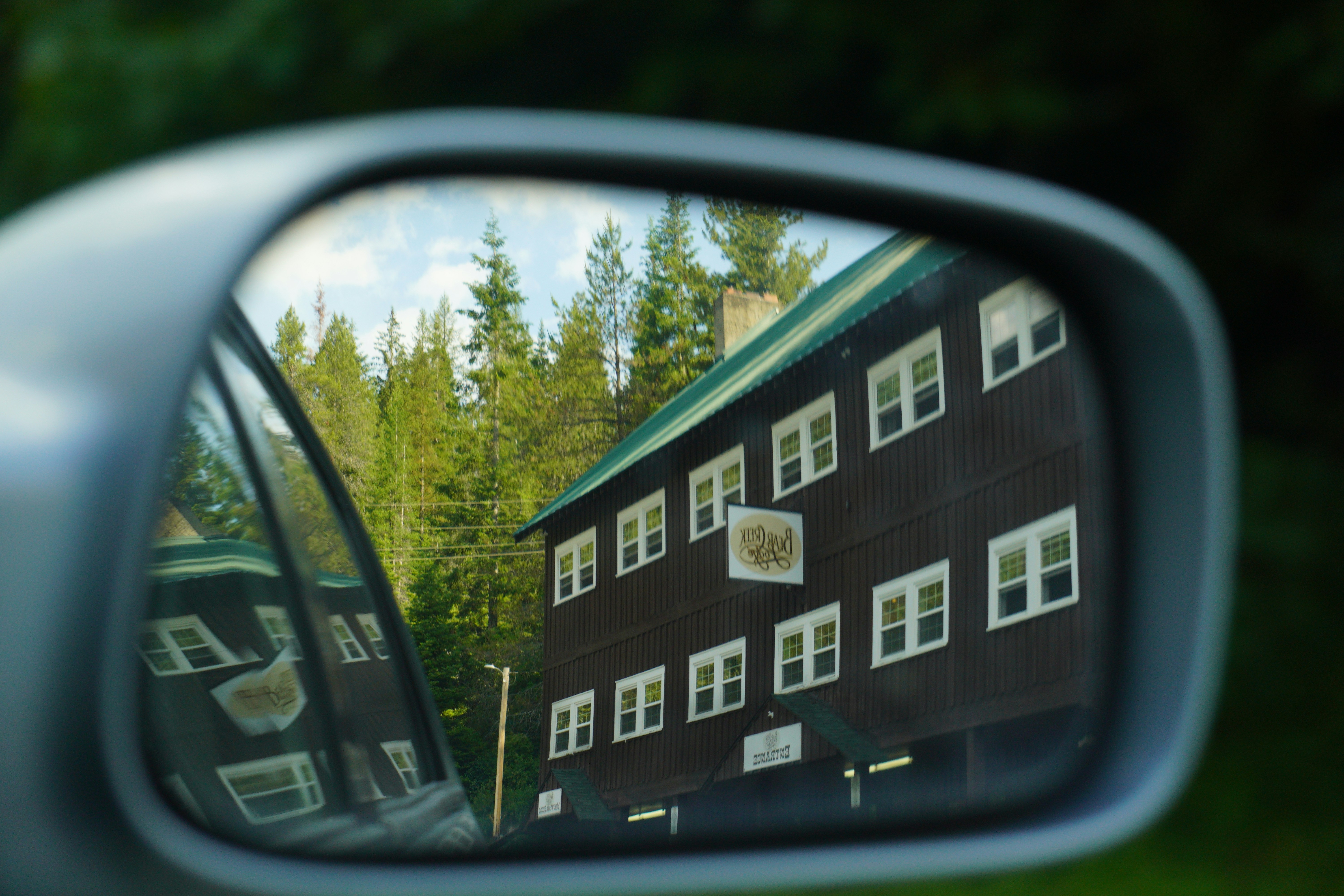 Mirror-Frame Vista: A dark wooden inn reflected in a side-view mirror amid pines