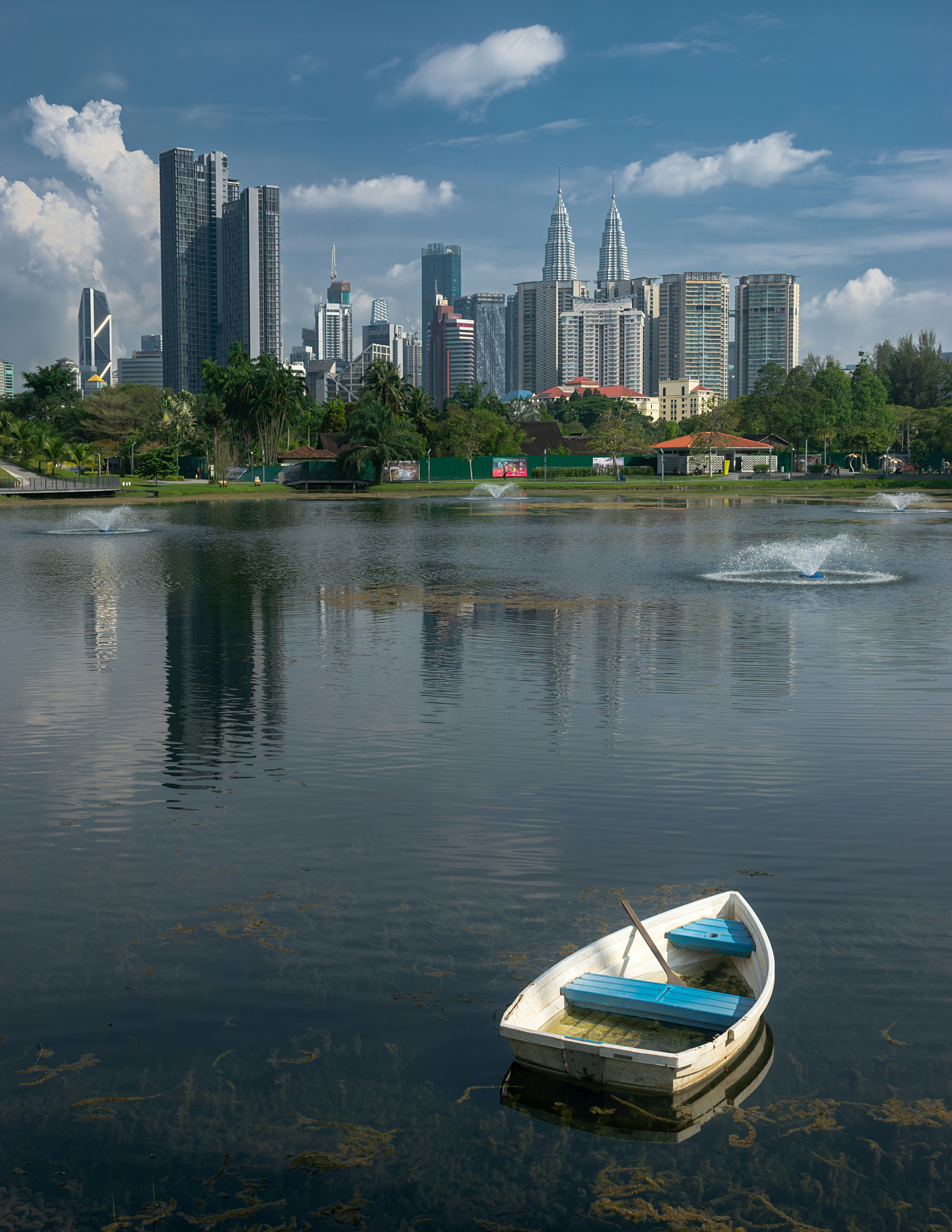 View of Kuala Lumpur skyline as seen from Tasik Titiwangsa on a blue sky day. Petronas Twin towers are on the right hand side. Malaysia, Apr/23. | a small boat floating on top of a lake