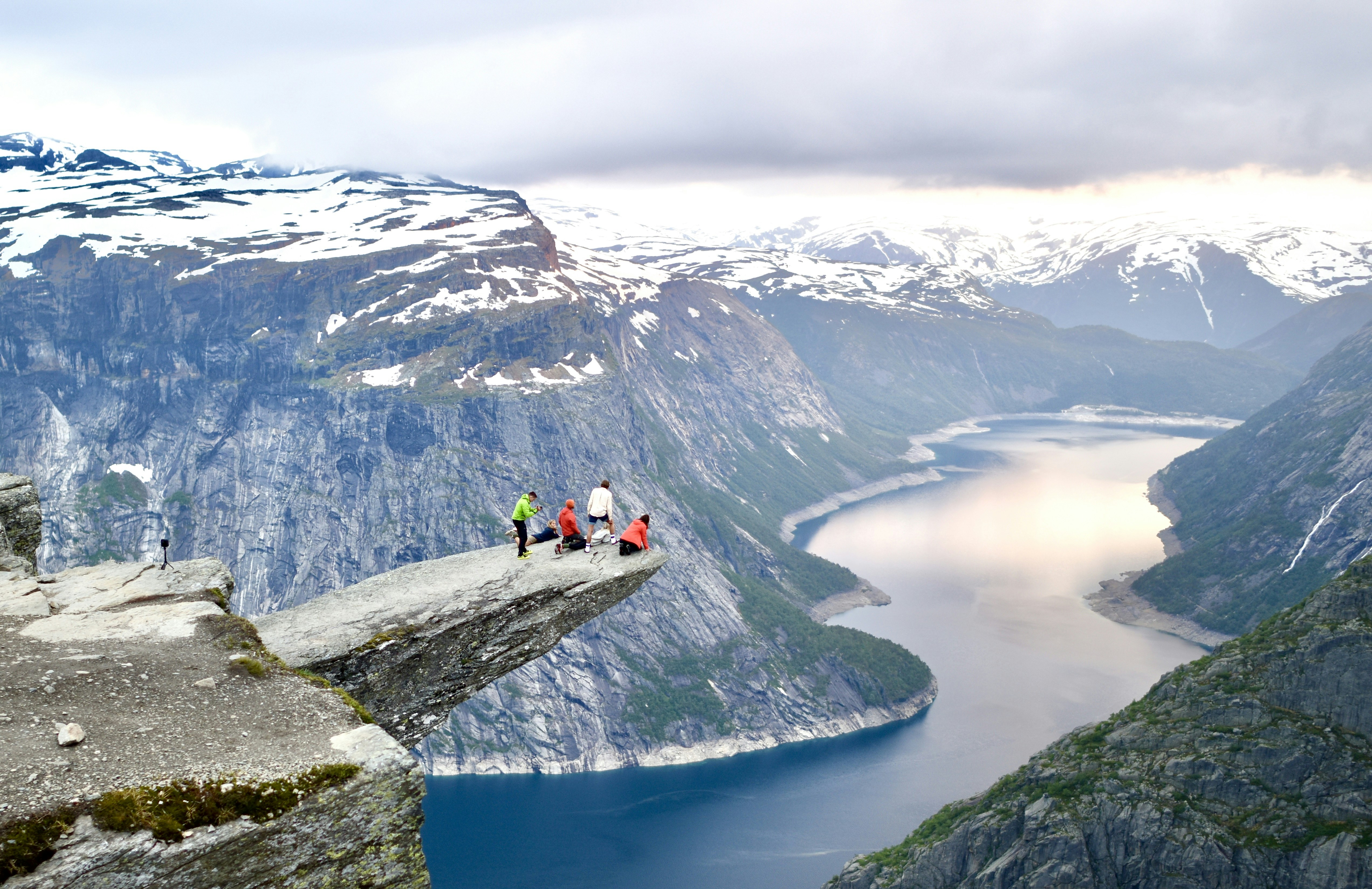 Hikers gather on the edge of Trolltunga overlooking a vast fjord and snowy mountains.