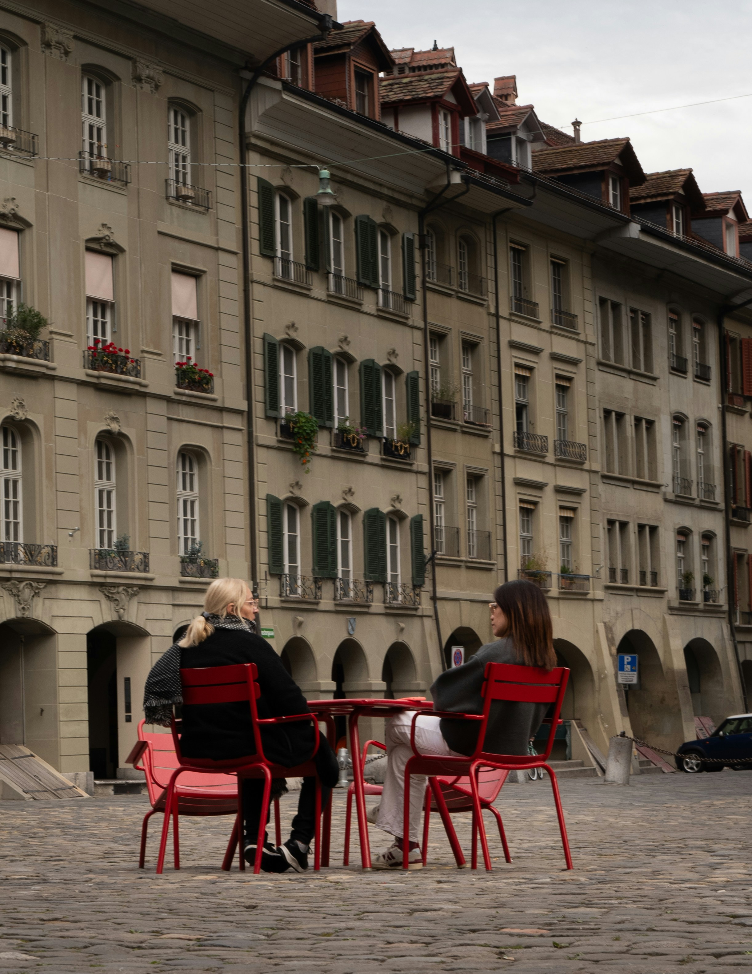 Two women sit at a small red-table café on a cobblestone street, facing each other. Historic European façades line the background, lending a timeless urban setting.