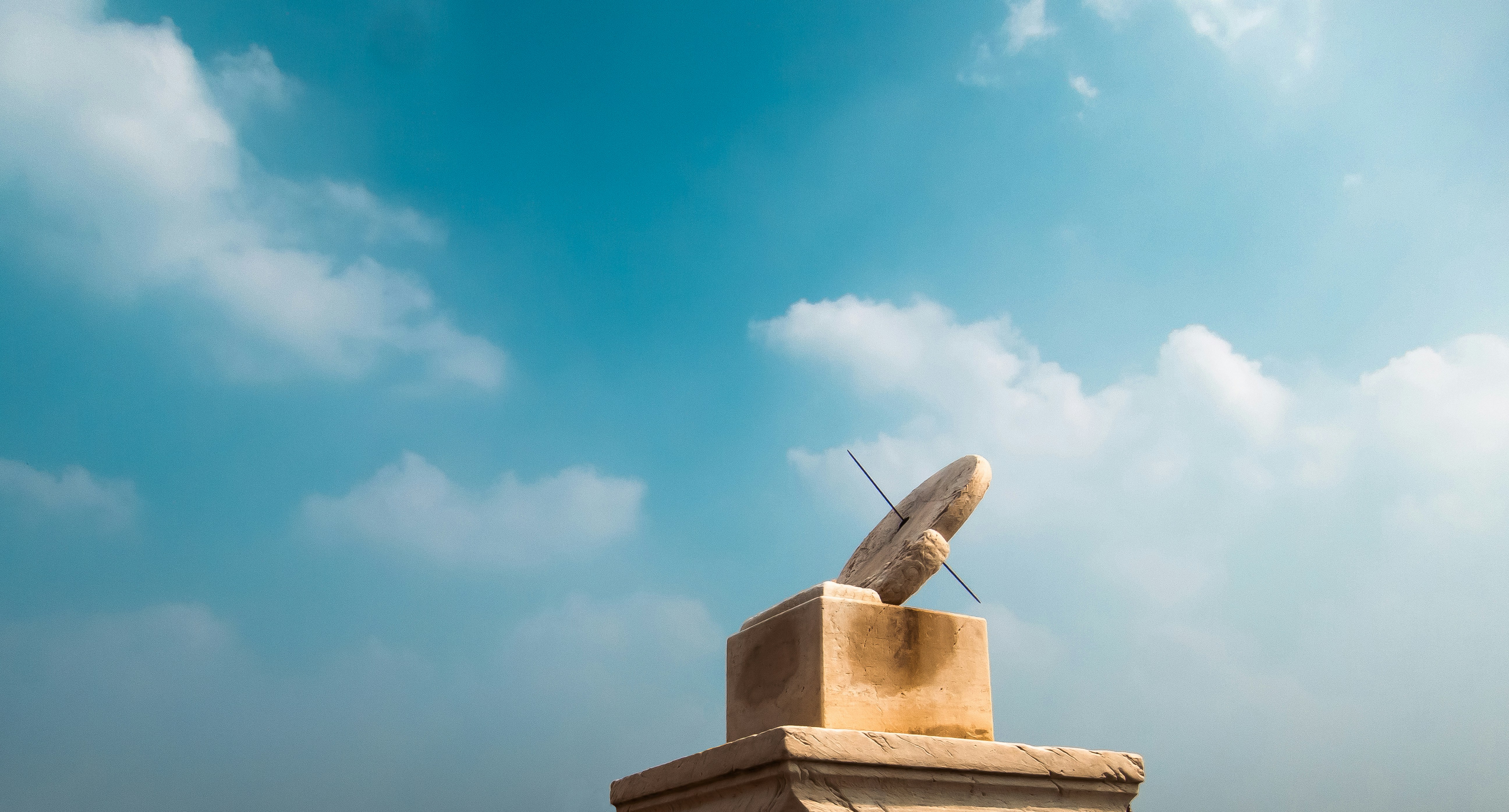 A sundial perched atop a stone pedestal, set against a backdrop of soft, billowing clouds. The delicate shadows cast by the gnomon indicate the passage of time.