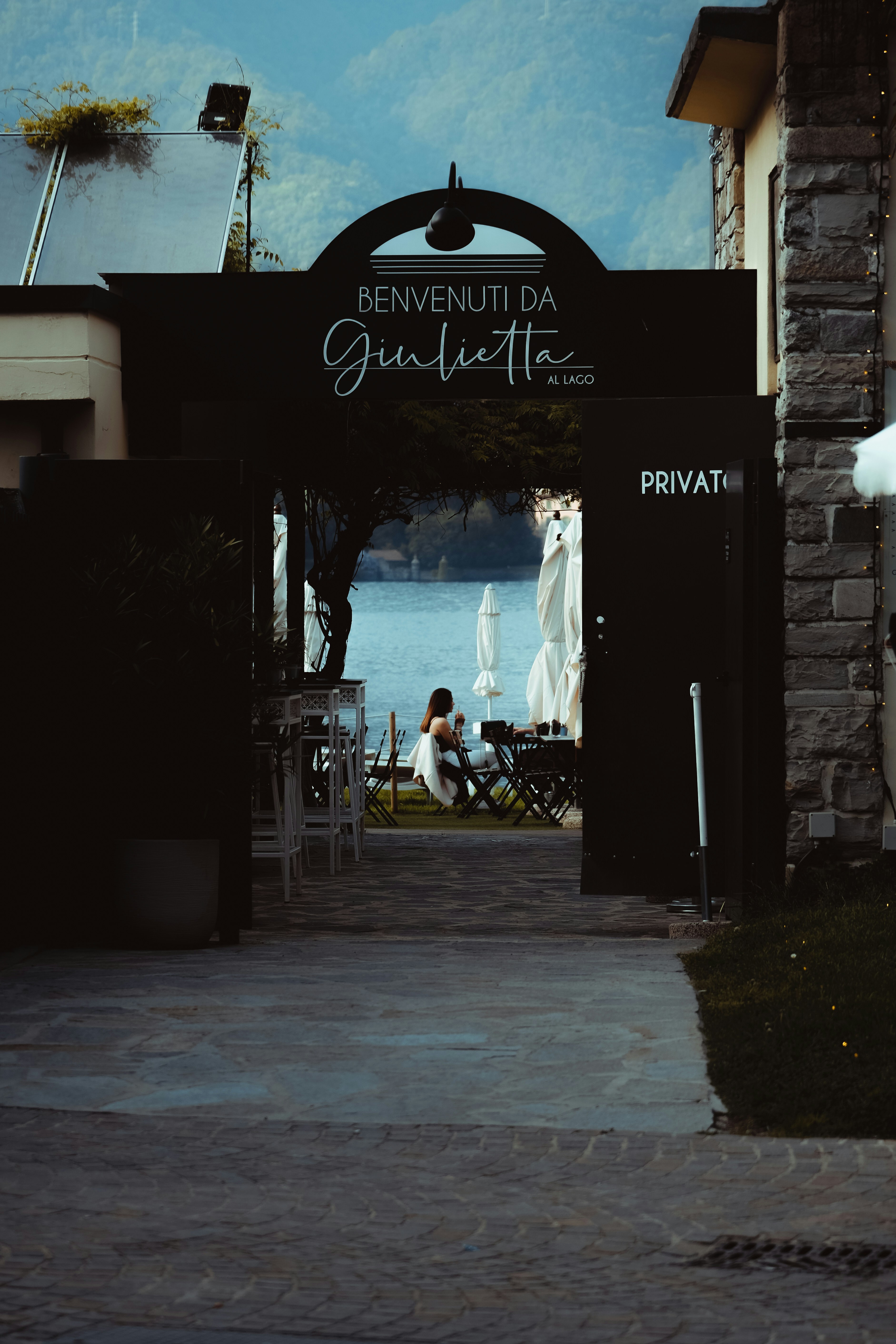 Charming entrance to a lakeside café with a welcoming sign and outdoor seating, framed by lush greenery. A patron enjoys the view in the background.