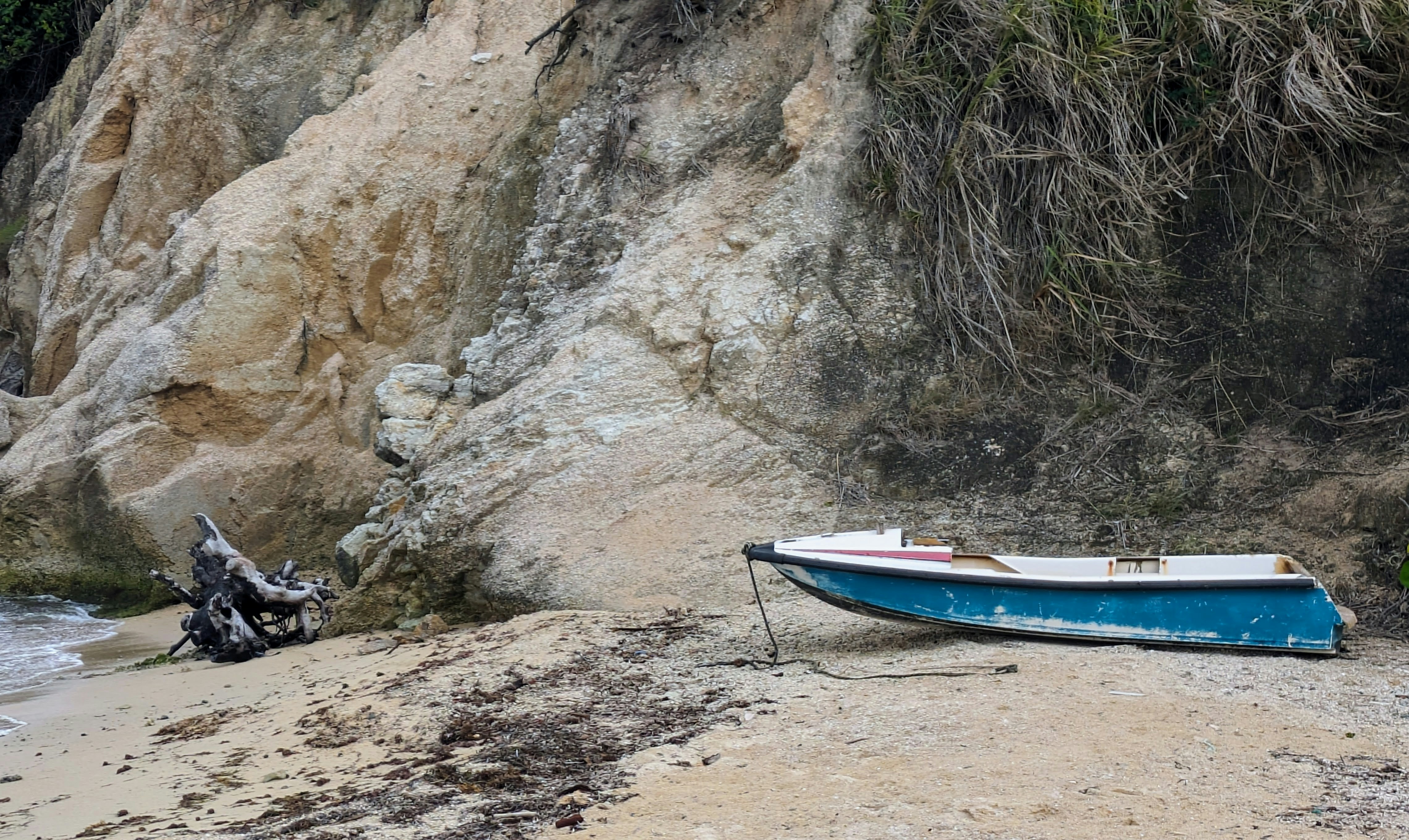 A blue rowboat rests on a sandy beach beside rugged cliffs and driftwood, with calm water in the background.