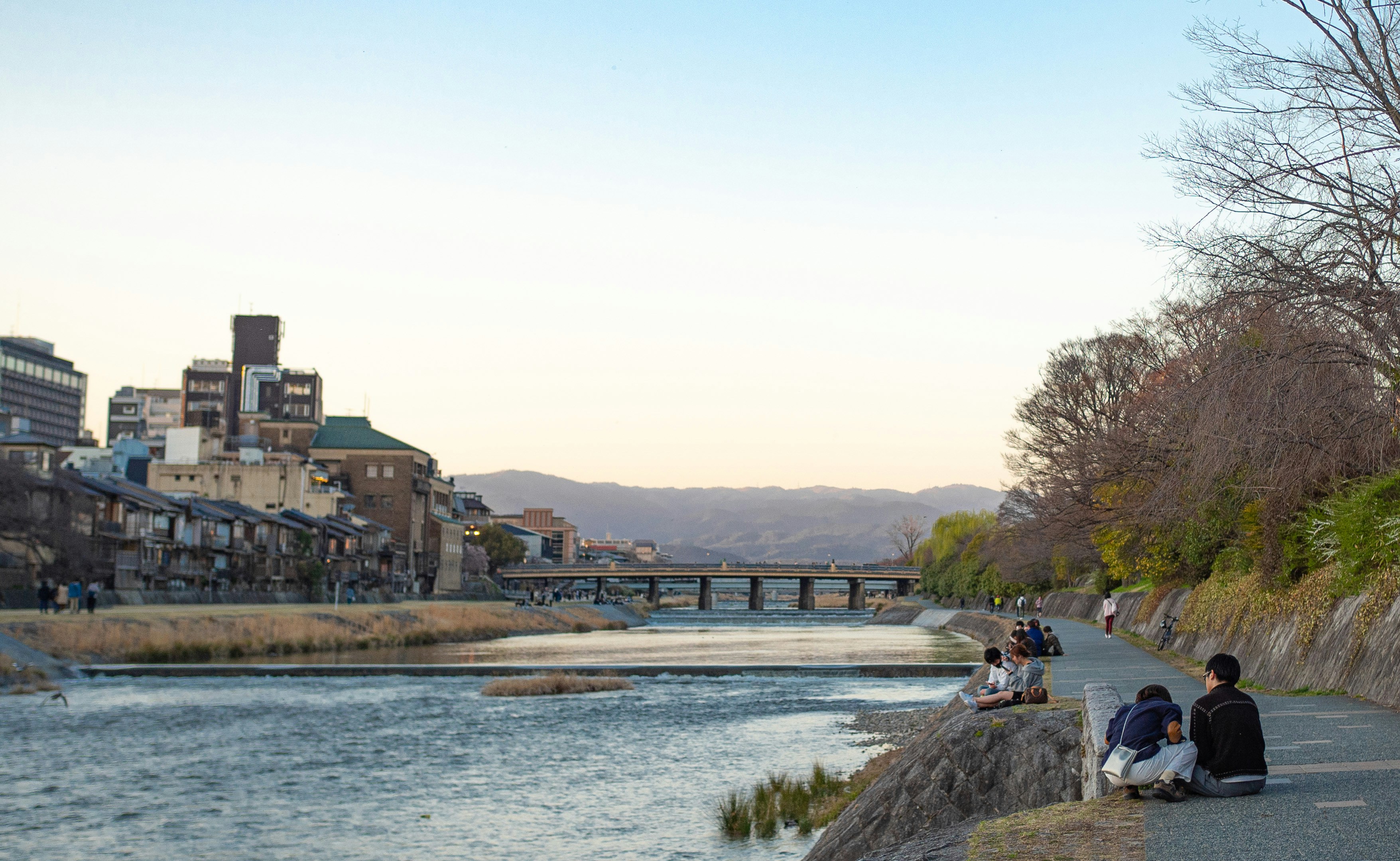 People relax along a riverbank with a cityscape and distant mountains under a clear evening sky.