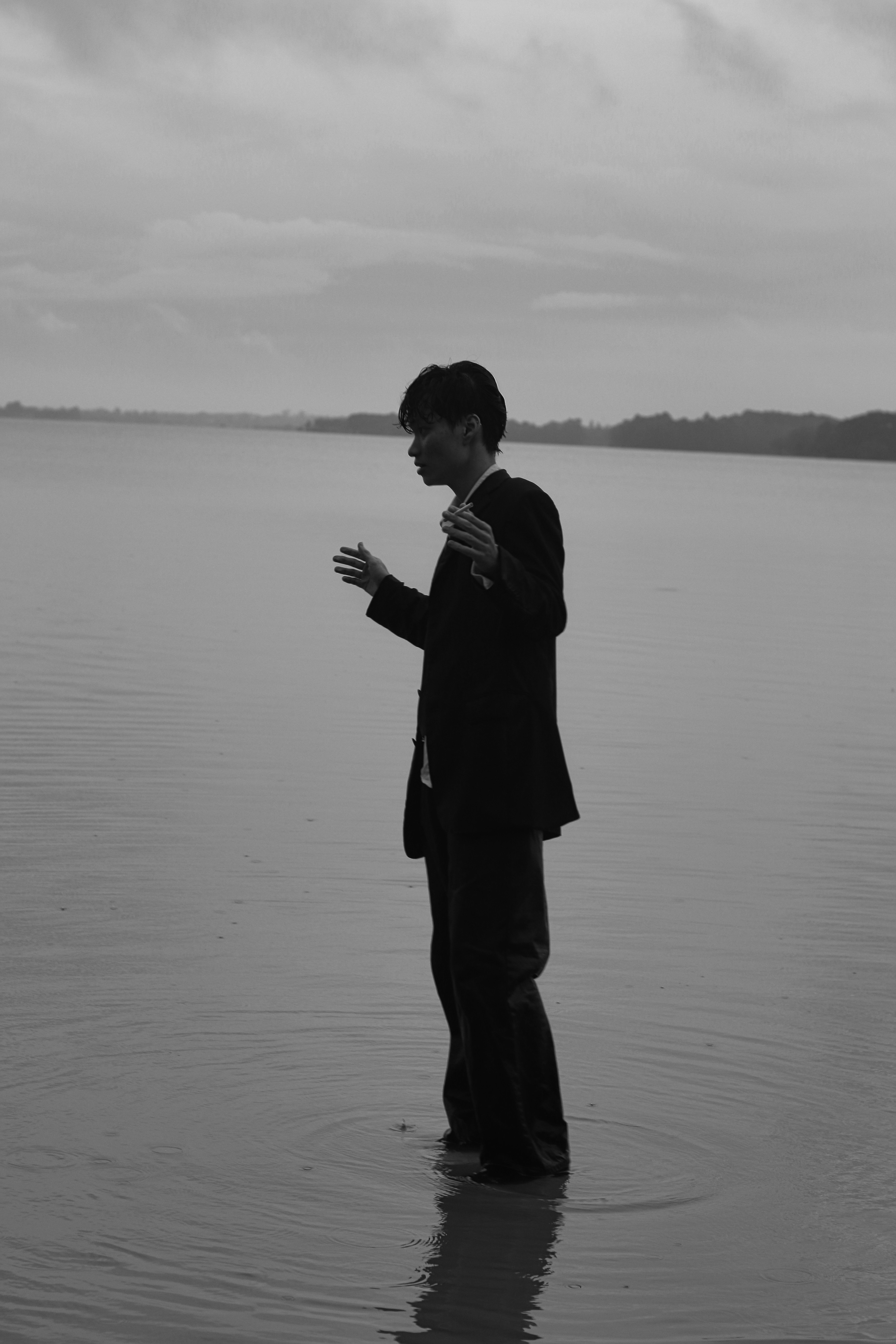 Person standing in shallow water, gesturing thoughtfully against a moody, overcast sky. The monochrome tones enhance the introspective atmosphere.