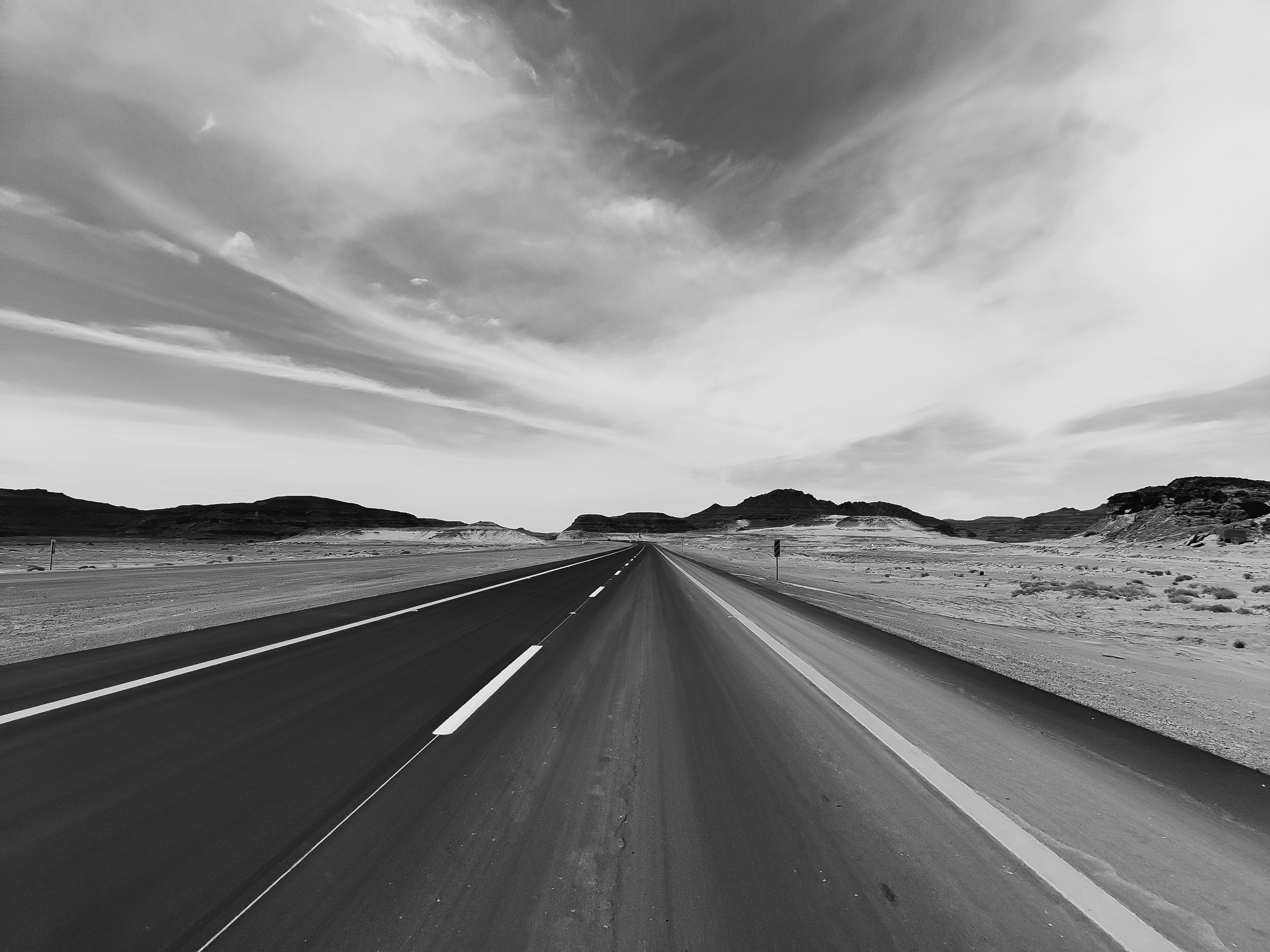 Long stretch of asphalt road cutting through barren desert landscape under a vast, cloudy sky.