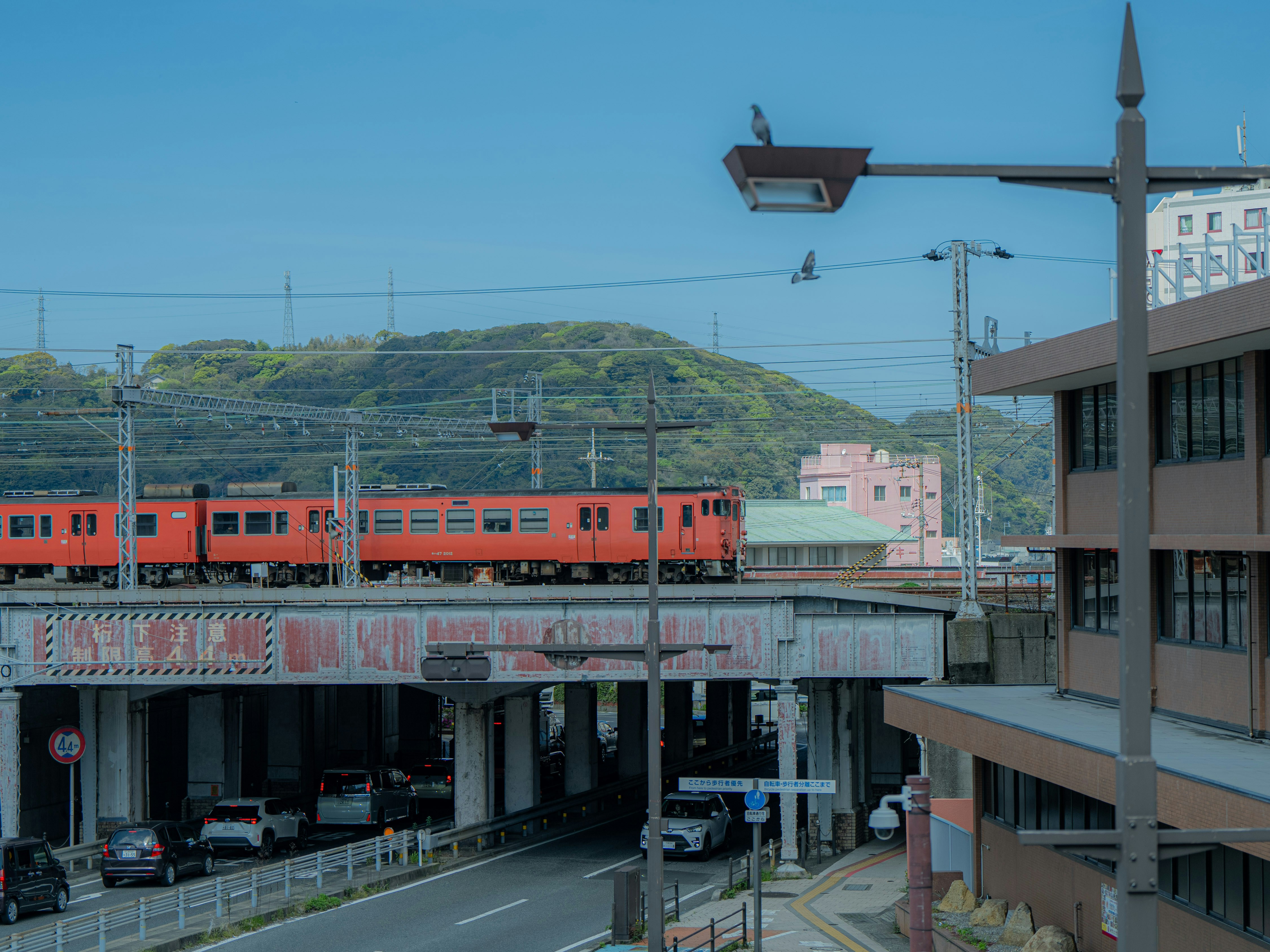 Red train crossing an elevated bridge near a modern building with a green hillside in the background.