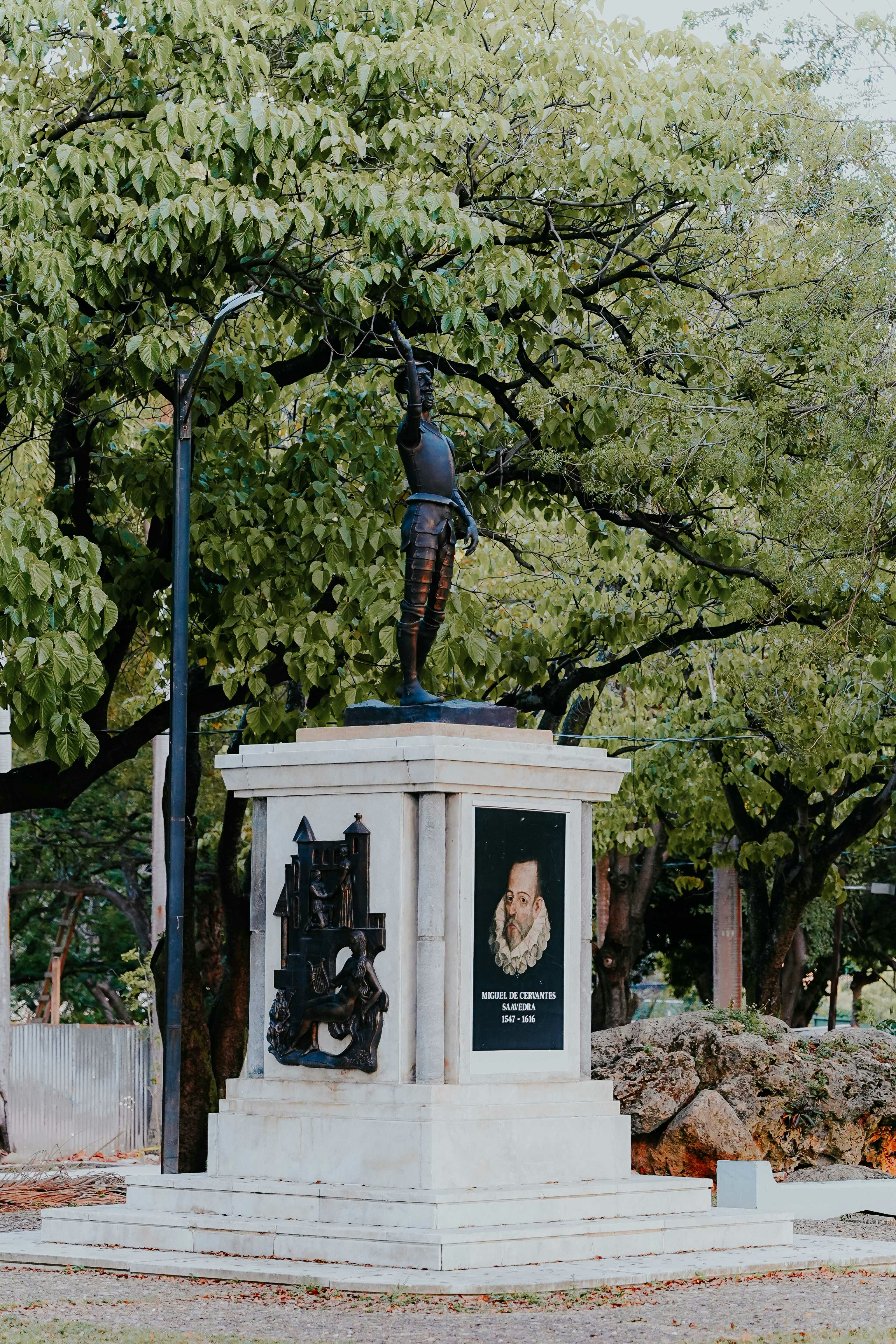 Statue pedestal crowned by a bronze figure beneath leafy canopy