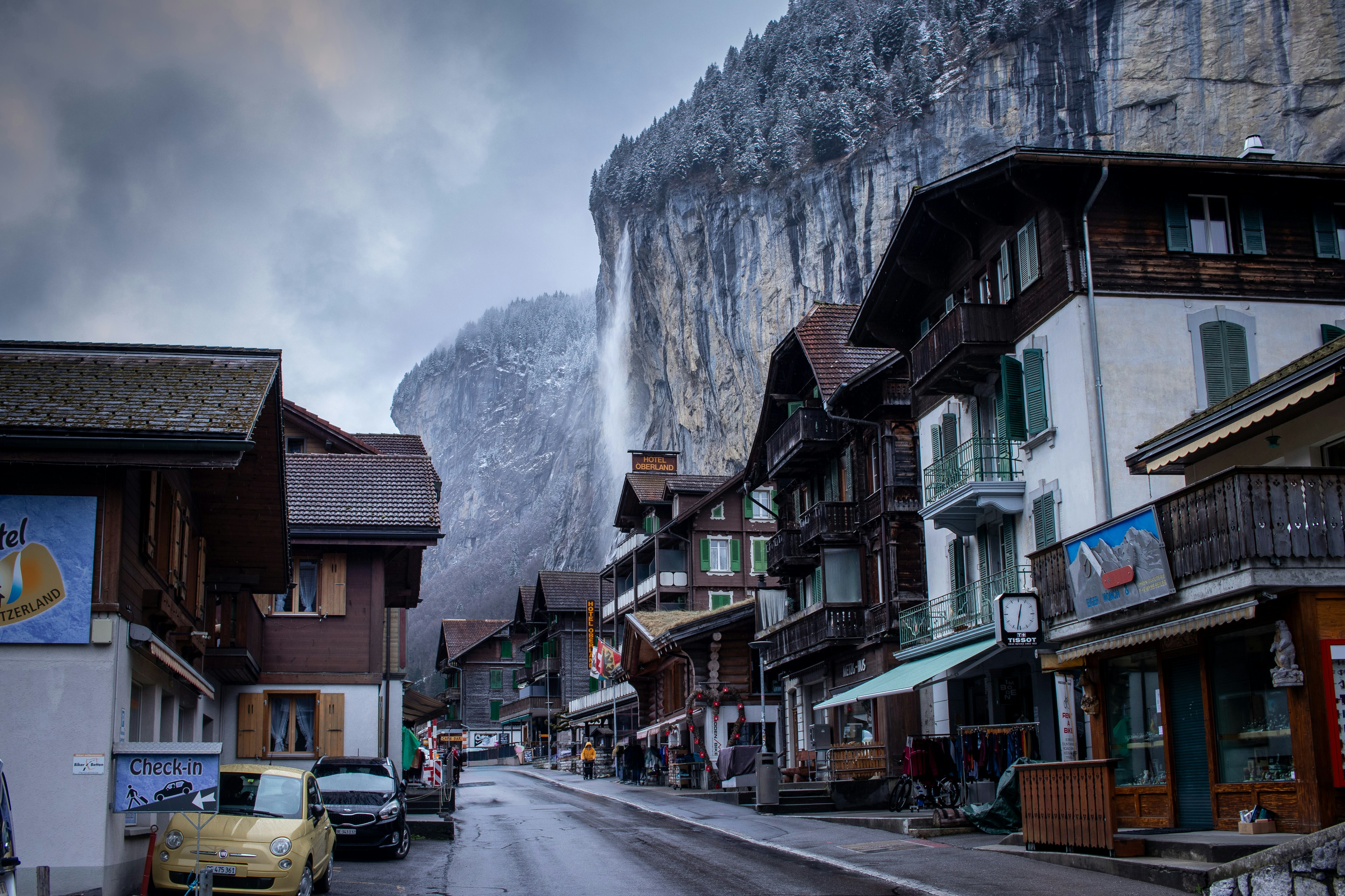 Quaint village street lined with traditional wooden chalets, set against towering cliffs and a misty sky. A sense of tranquility permeates the scene.