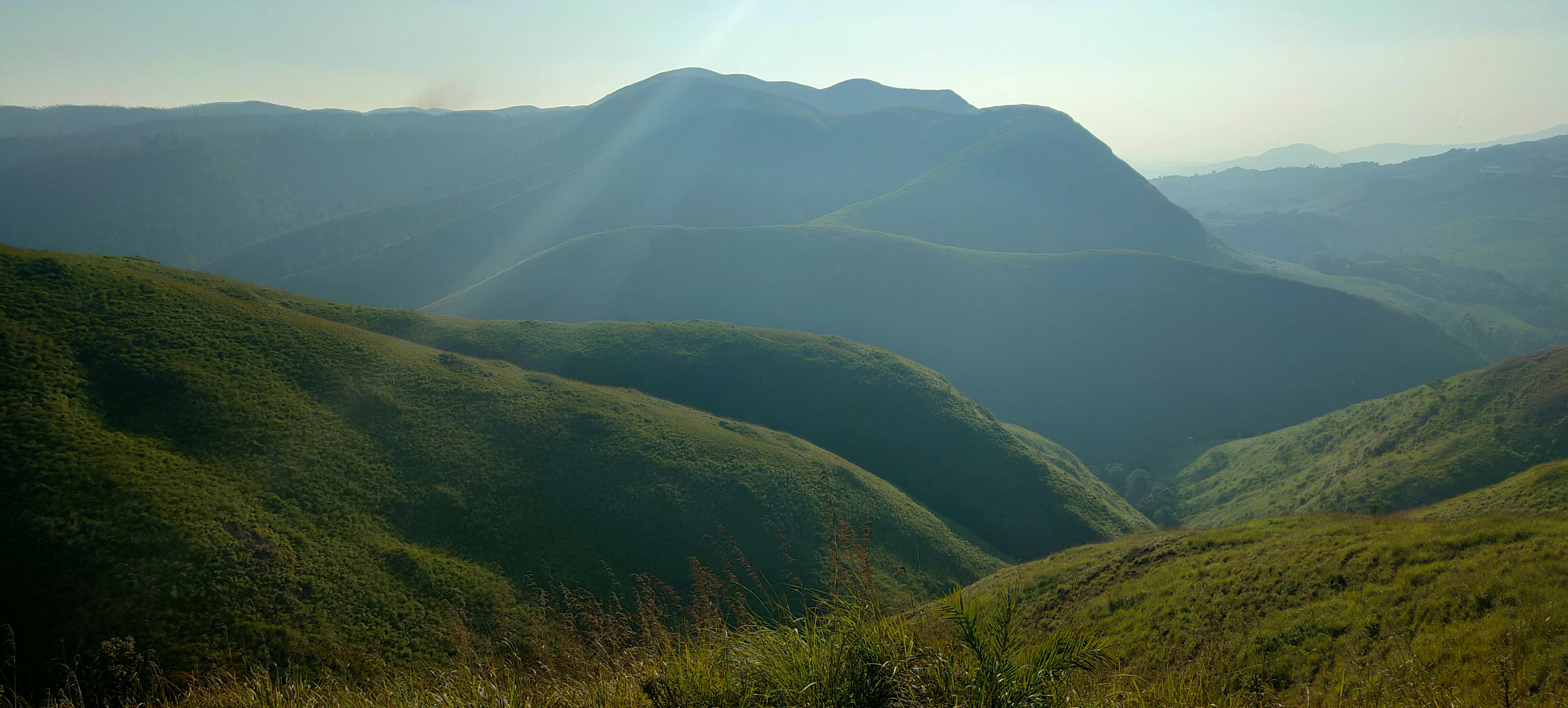 Landscape photograph of rolling green hills fading into distant ridges under soft morning light. Sunbeams pierce the haze, highlighting the textured valley contours.